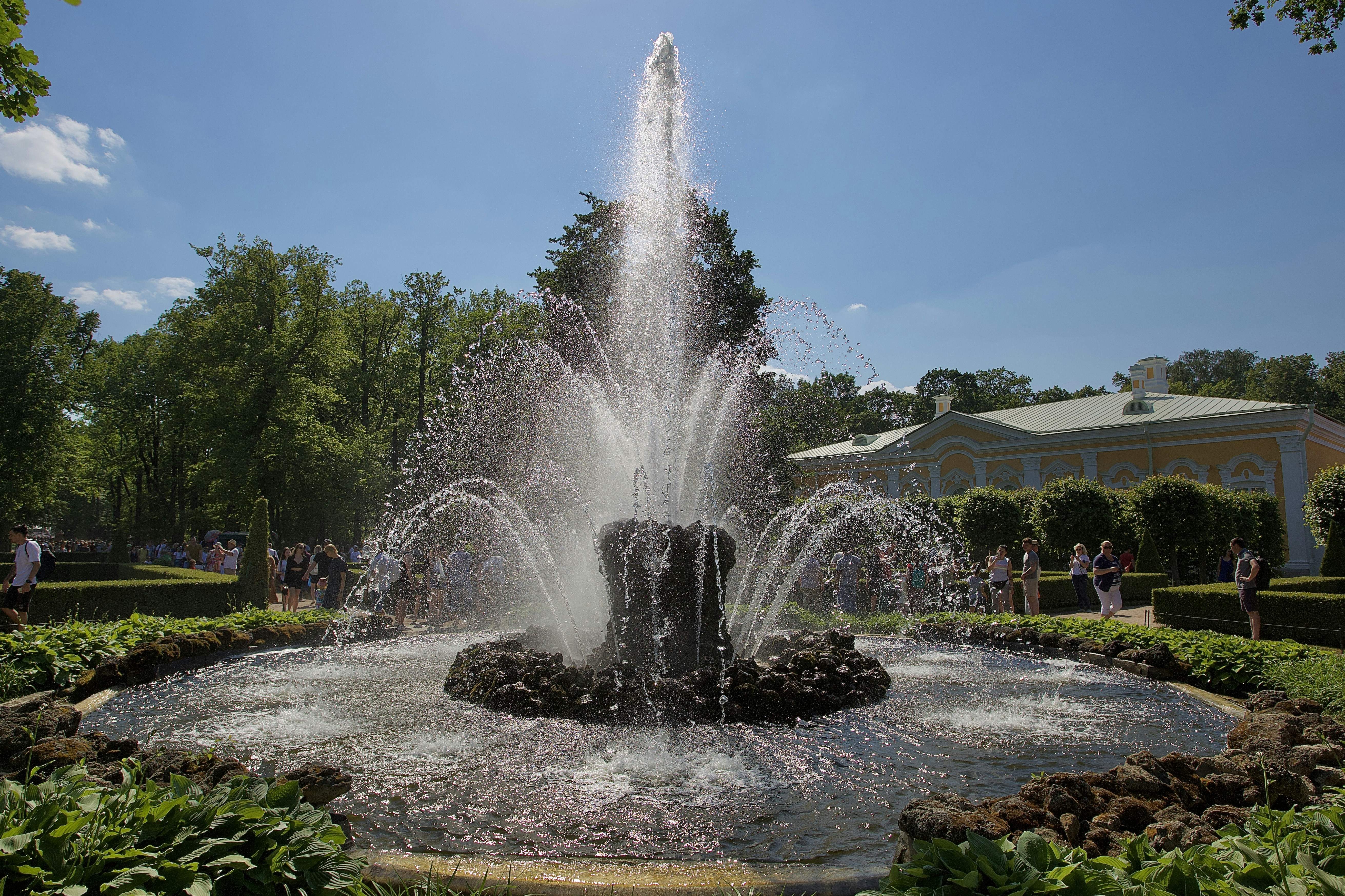 Majestic fountain surrounded by lush greenery and a historic building, showcasing a vibrant display of water jets under a clear blue sky.