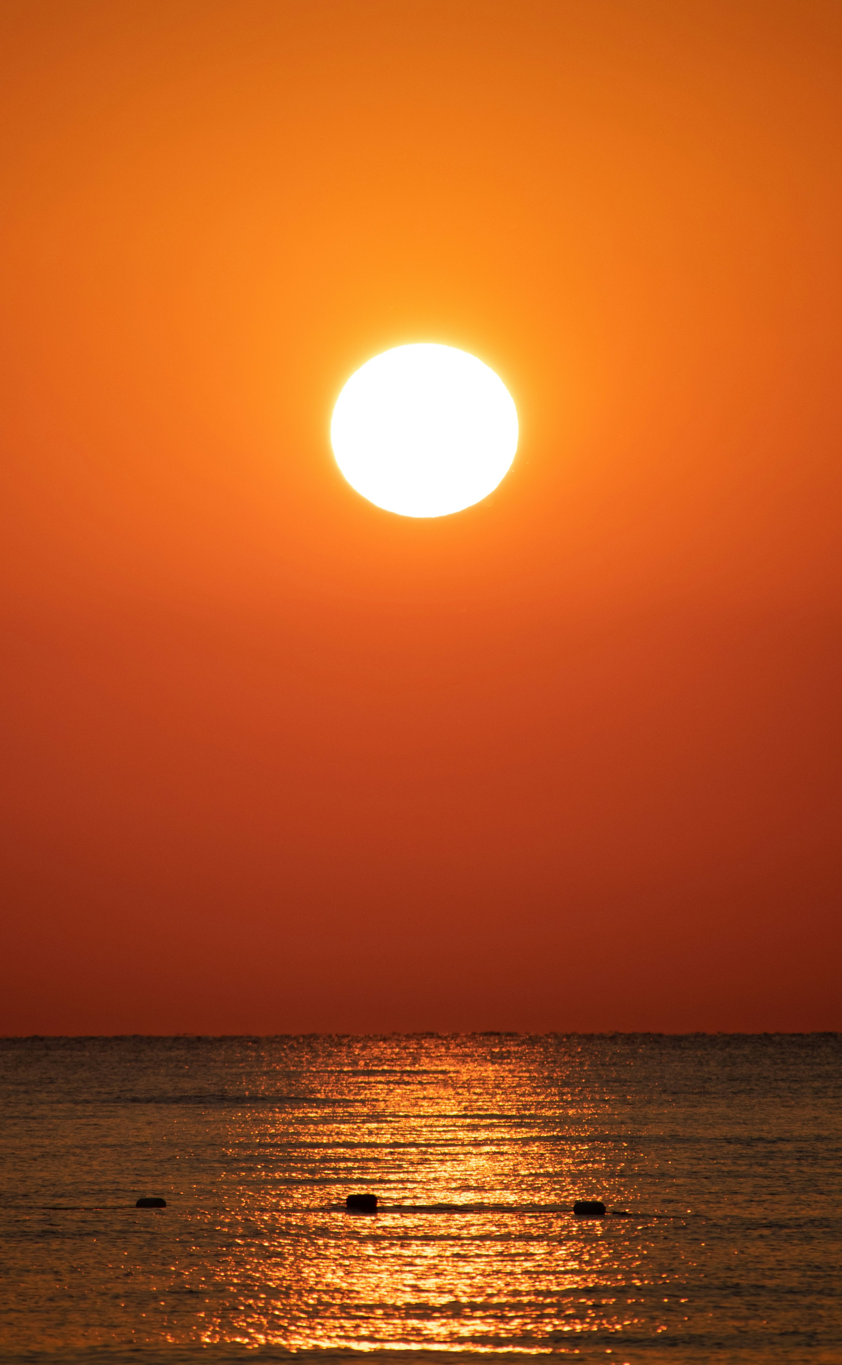 Bright sun setting over a calm sea, casting reflections on the water's surface. The warm orange gradient fills the sky.