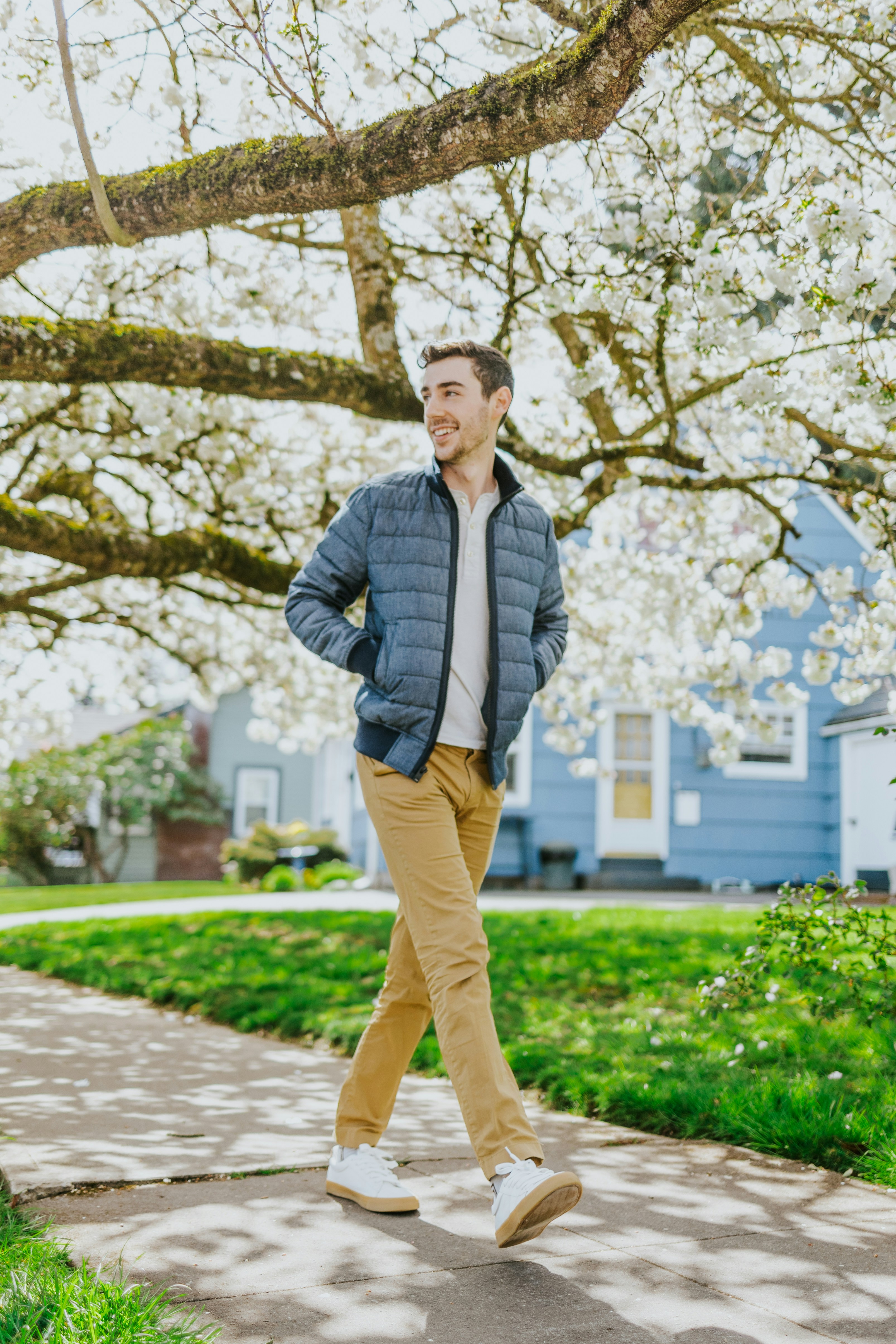 Man walking along a sidewalk beneath a flowering tree, exuding a relaxed spring vibe. The background features a charming blue house.