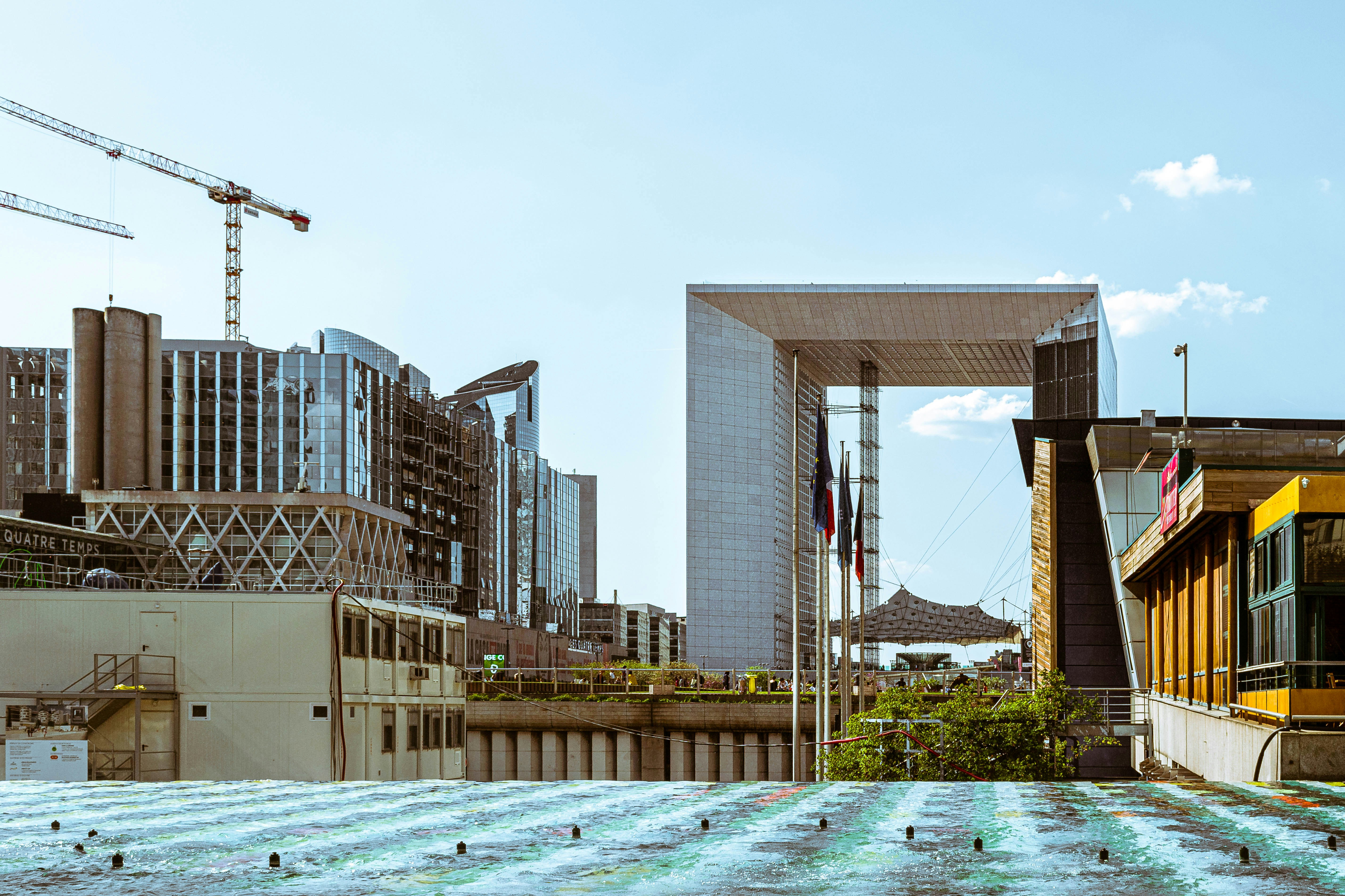 Modern white concrete archway framed by urban buildings and cranes under a clear blue sky.