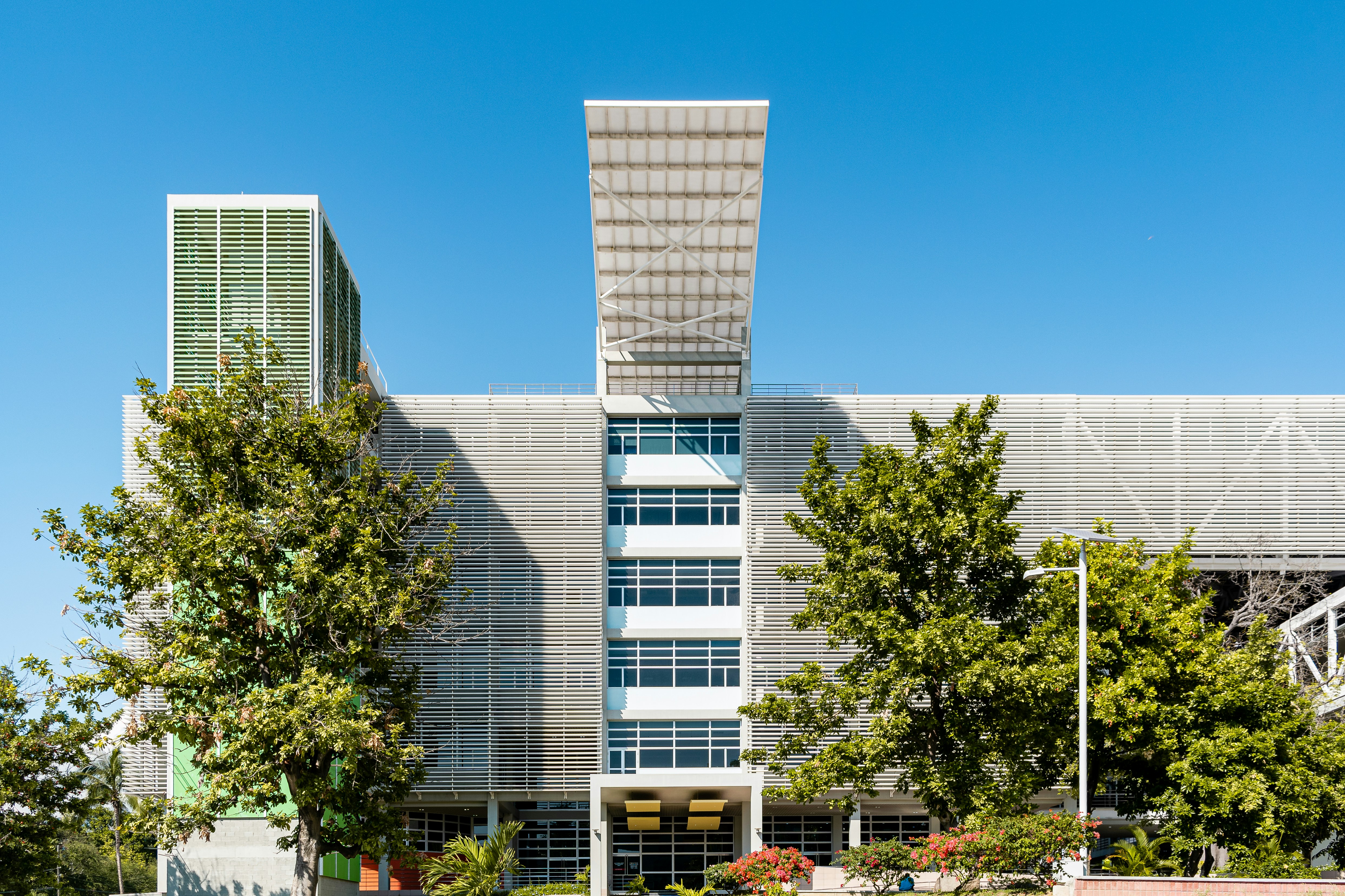 Contemporary building facade with geometric design elements surrounded by lush trees under a clear blue sky.