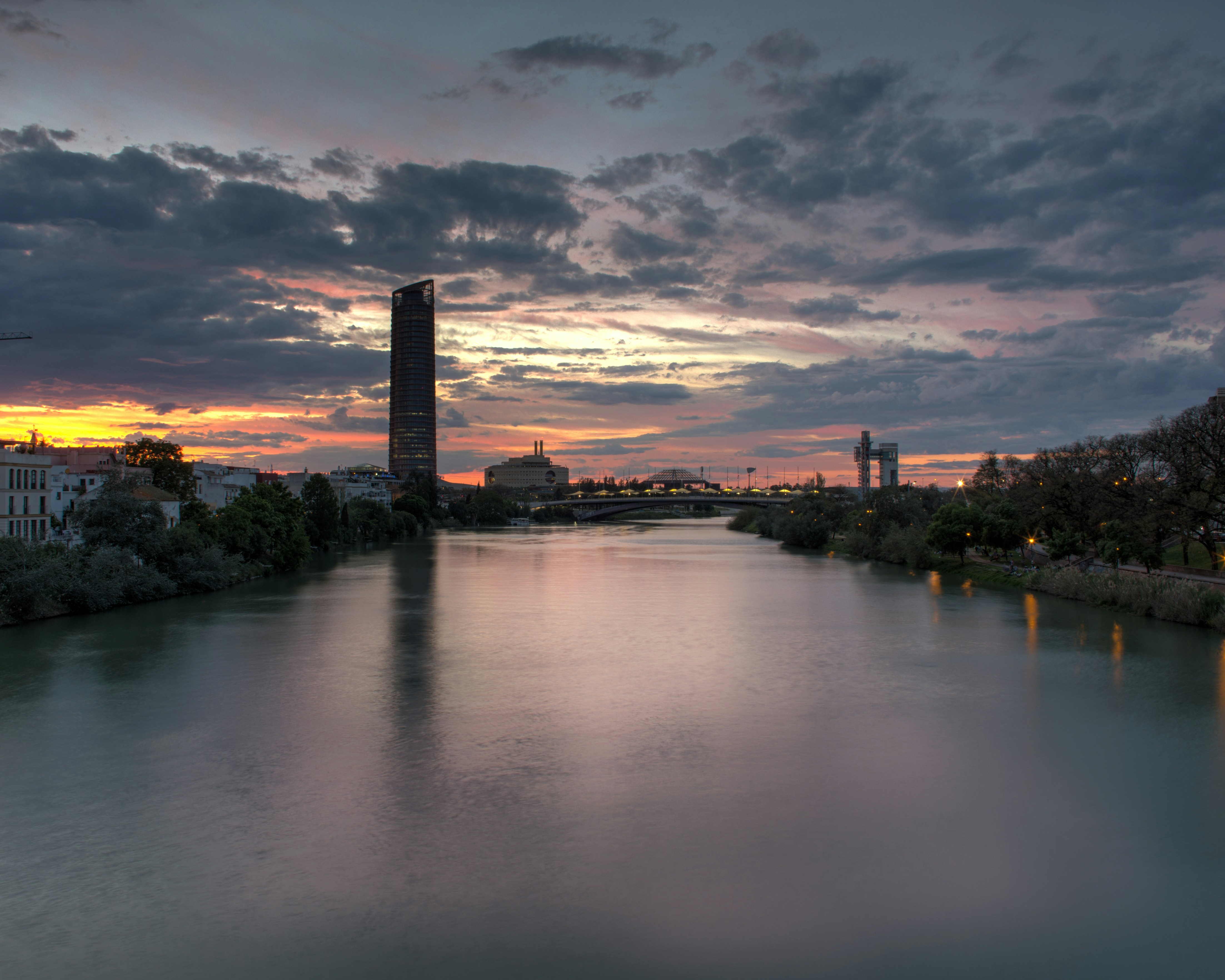 Calm river reflecting a dramatic sunset sky with silhouetted cityscape.