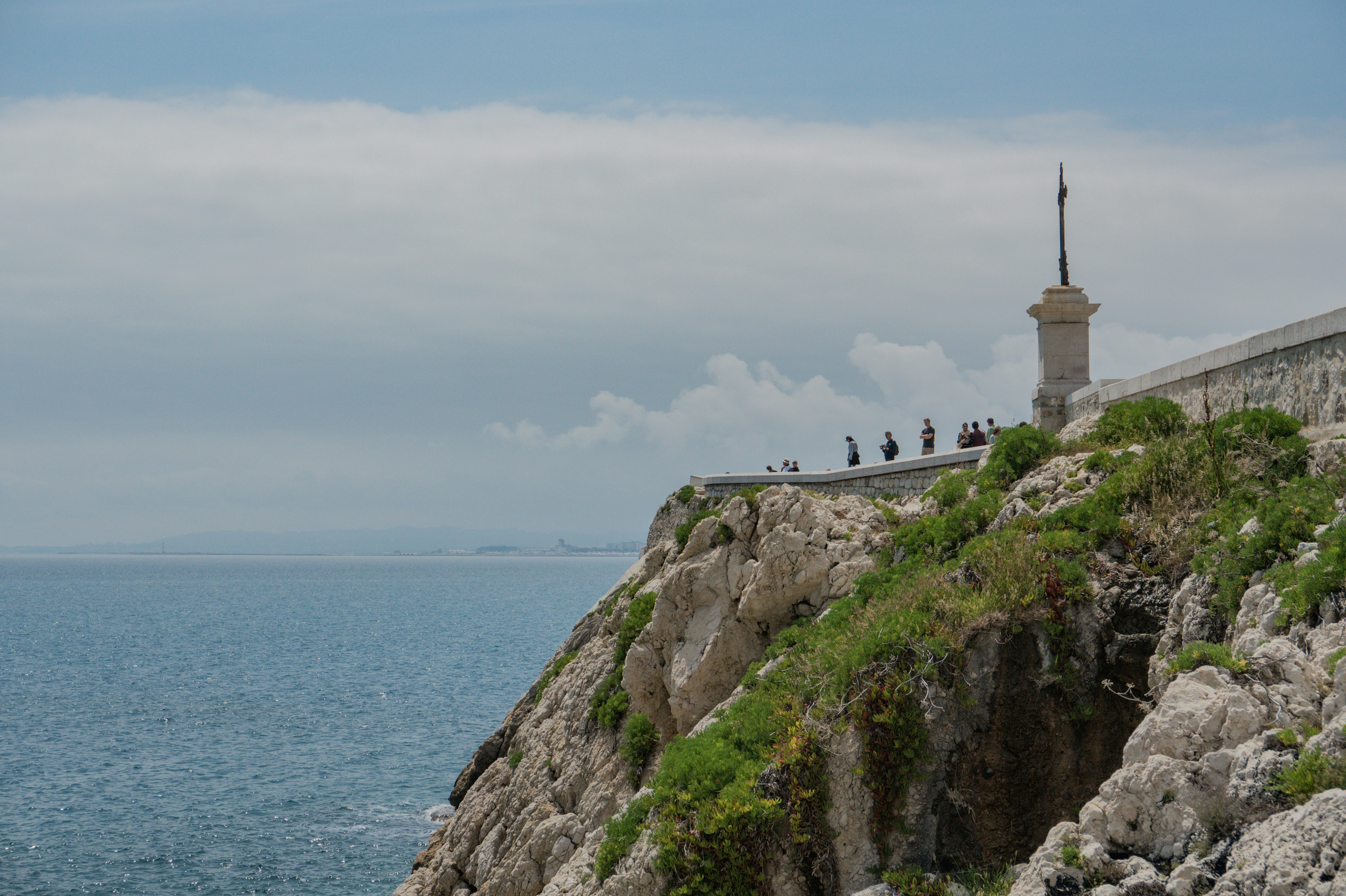 Visitors walking along a coastal path atop a rocky cliff, overlooking the serene ocean under a cloudy sky.