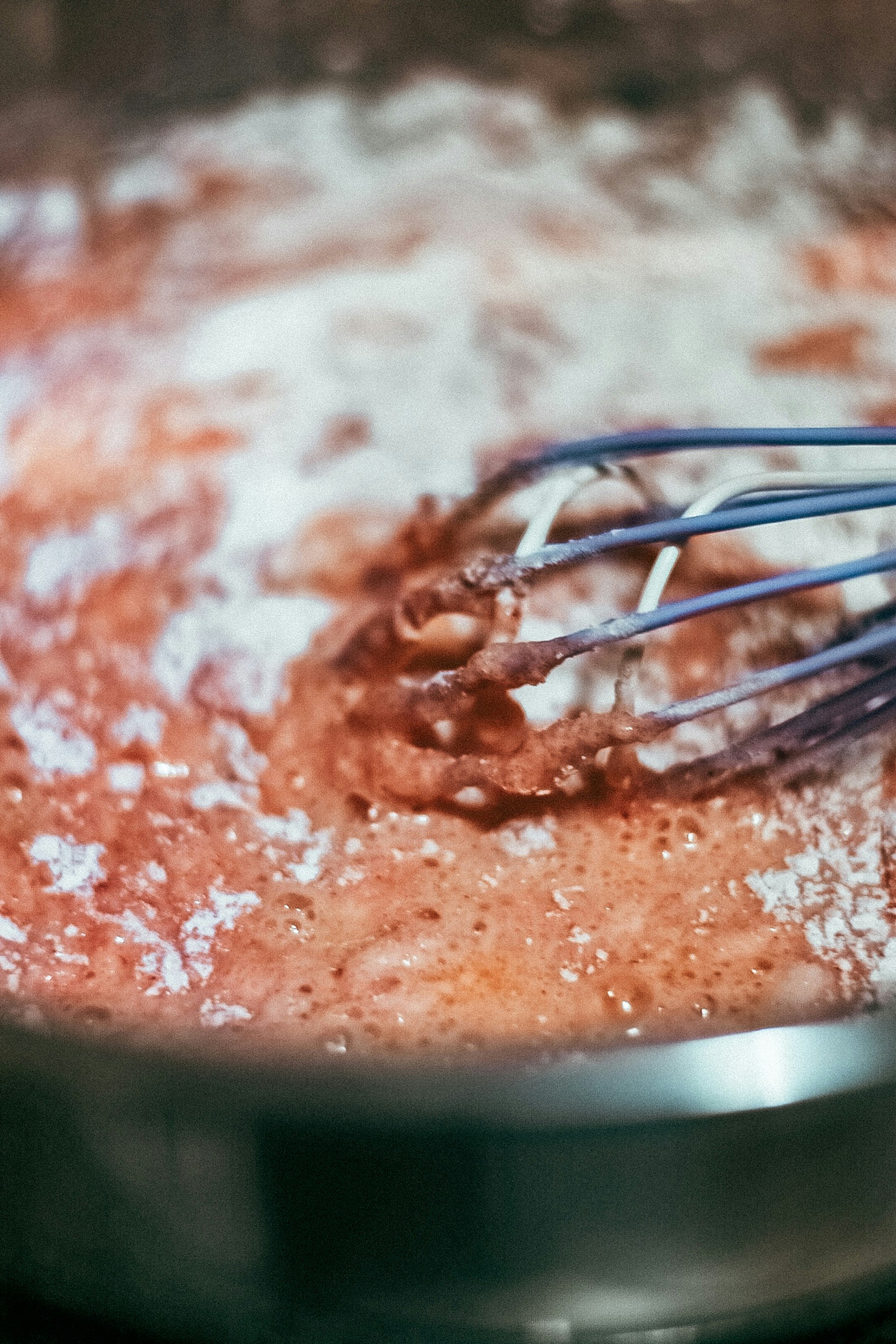 A close-up of a whisk mixing a bubbly, frothy mixture in a stainless steel bowl, with flour dusting the surface. The scene captures the essence of cooking in action.