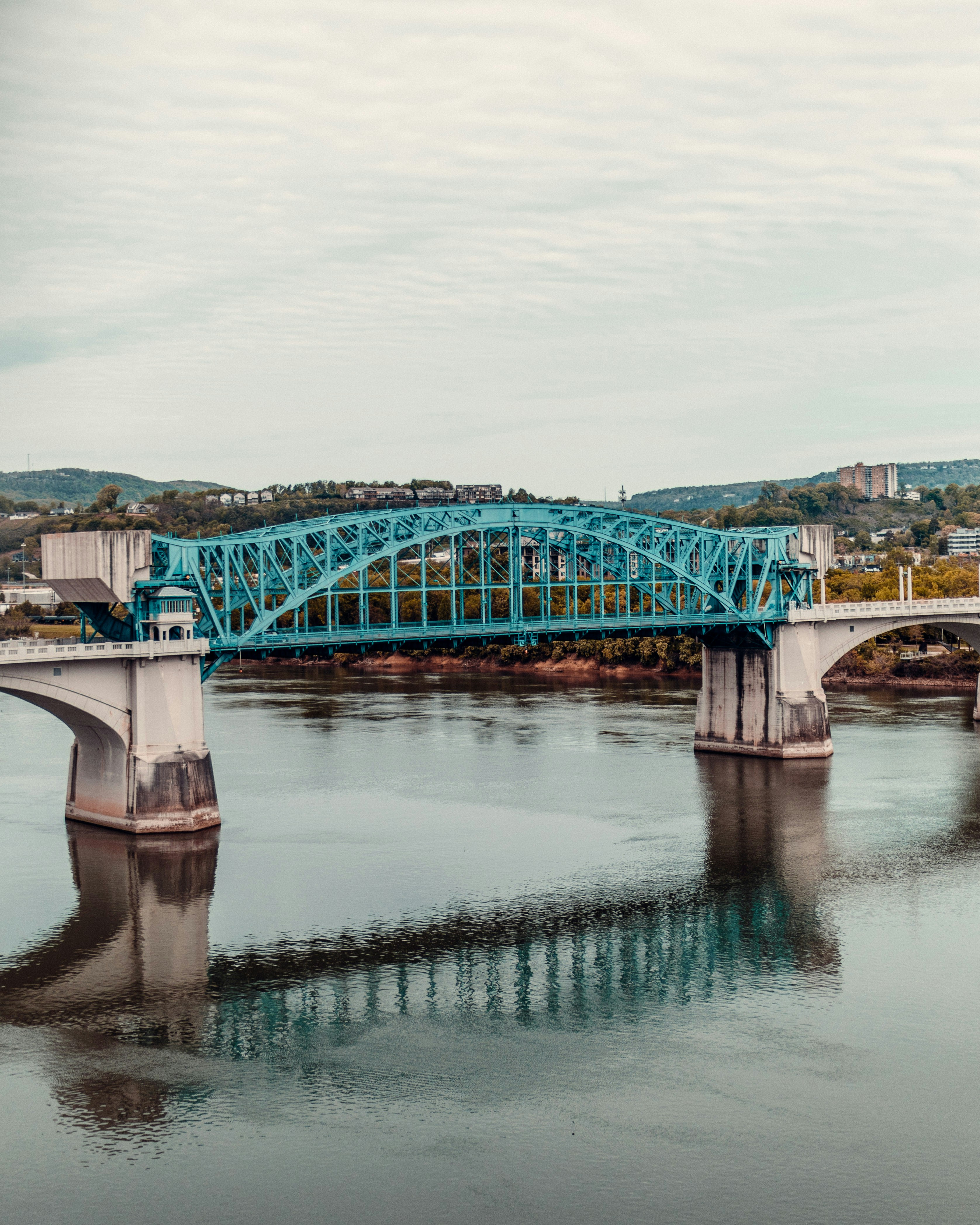 Vibrant turquoise bridge spanning a serene river, reflecting its colors on the water's surface under a cloudy sky.