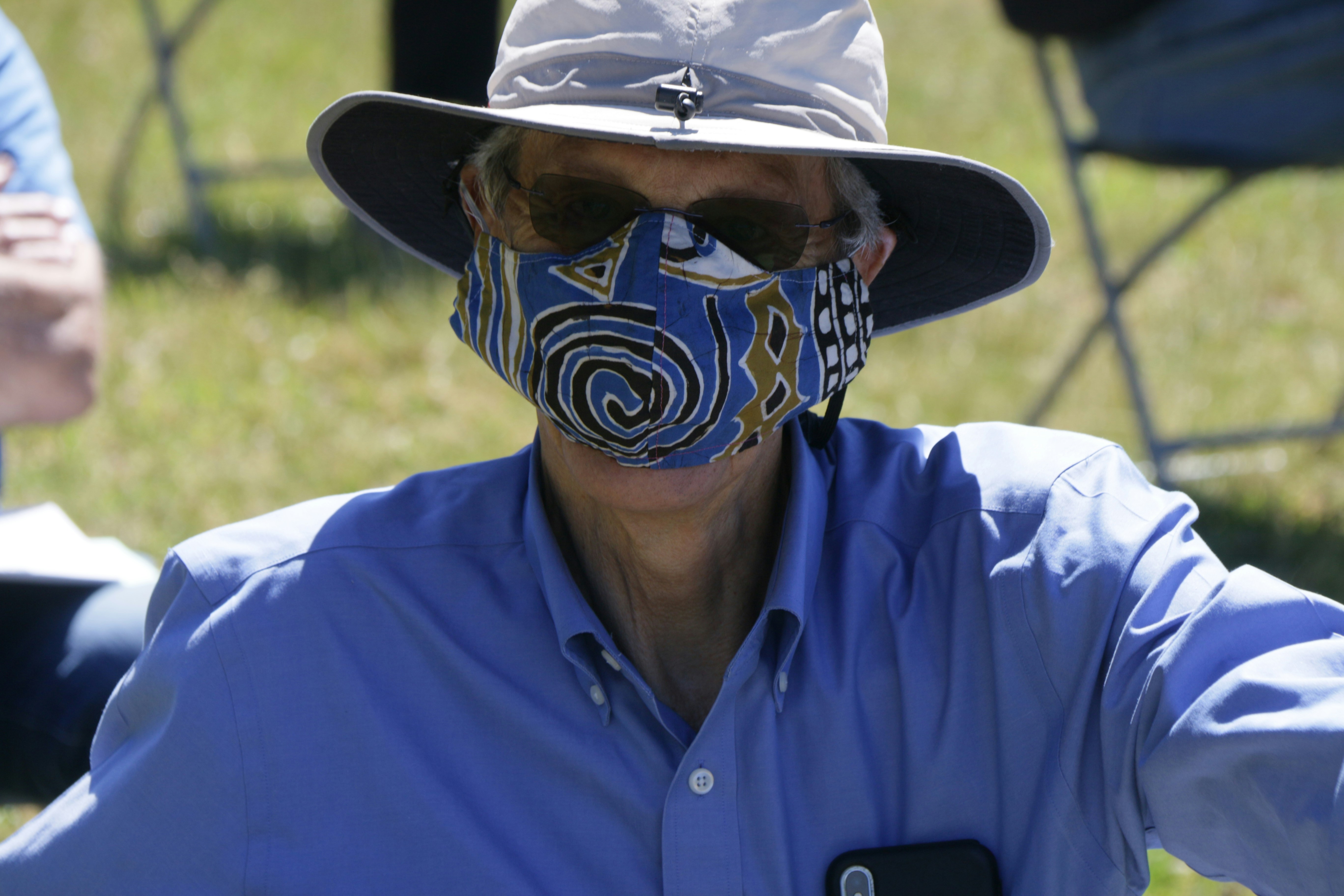 Individual wearing a patterned mask and wide-brimmed hat, seated outdoors in a grassy area. The scene captures a moment of social interaction amidst health precautions.