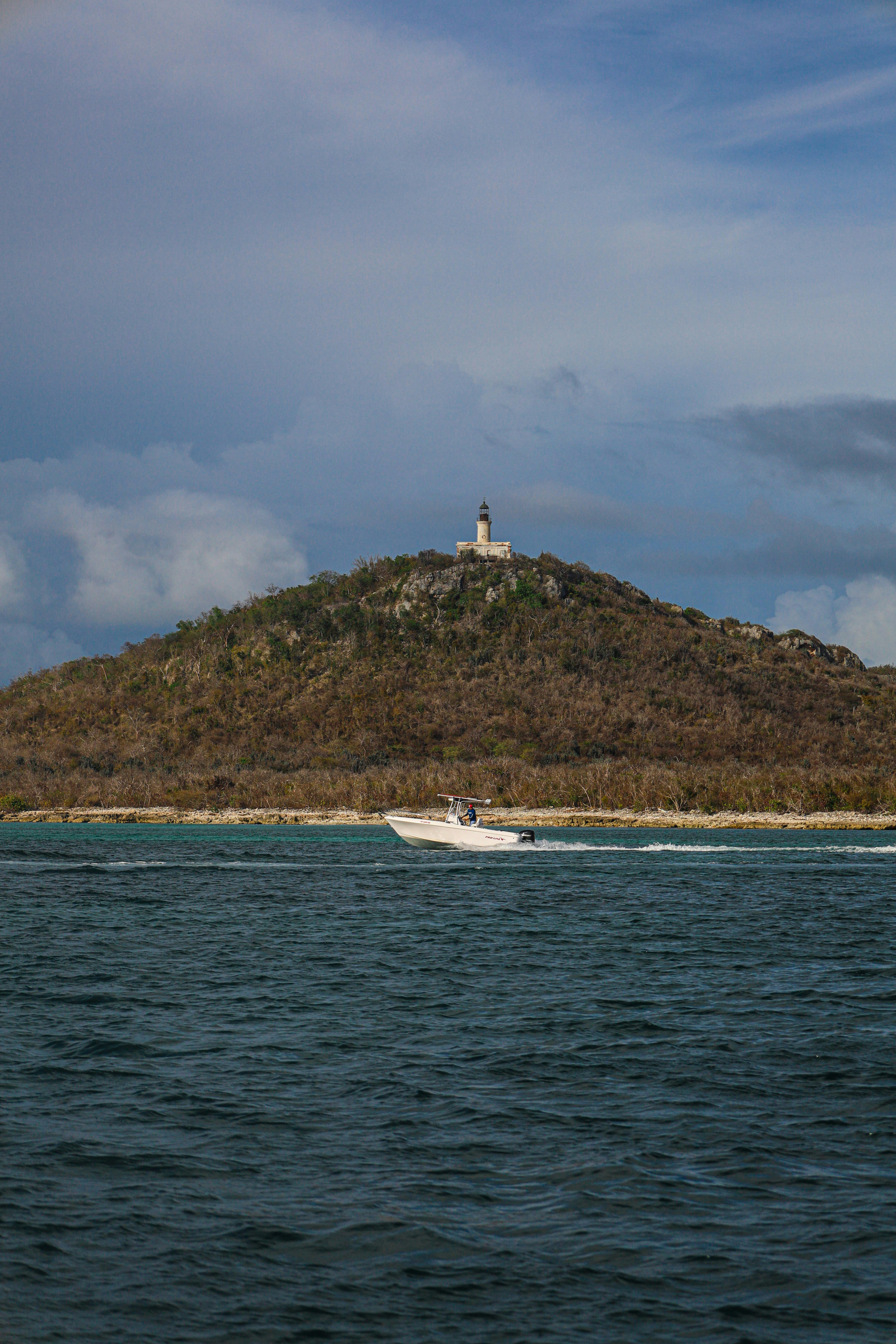 A speedboat glides across the water near a lush, hilly landscape topped with a lighthouse. The scene captures a tranquil moment on the water.