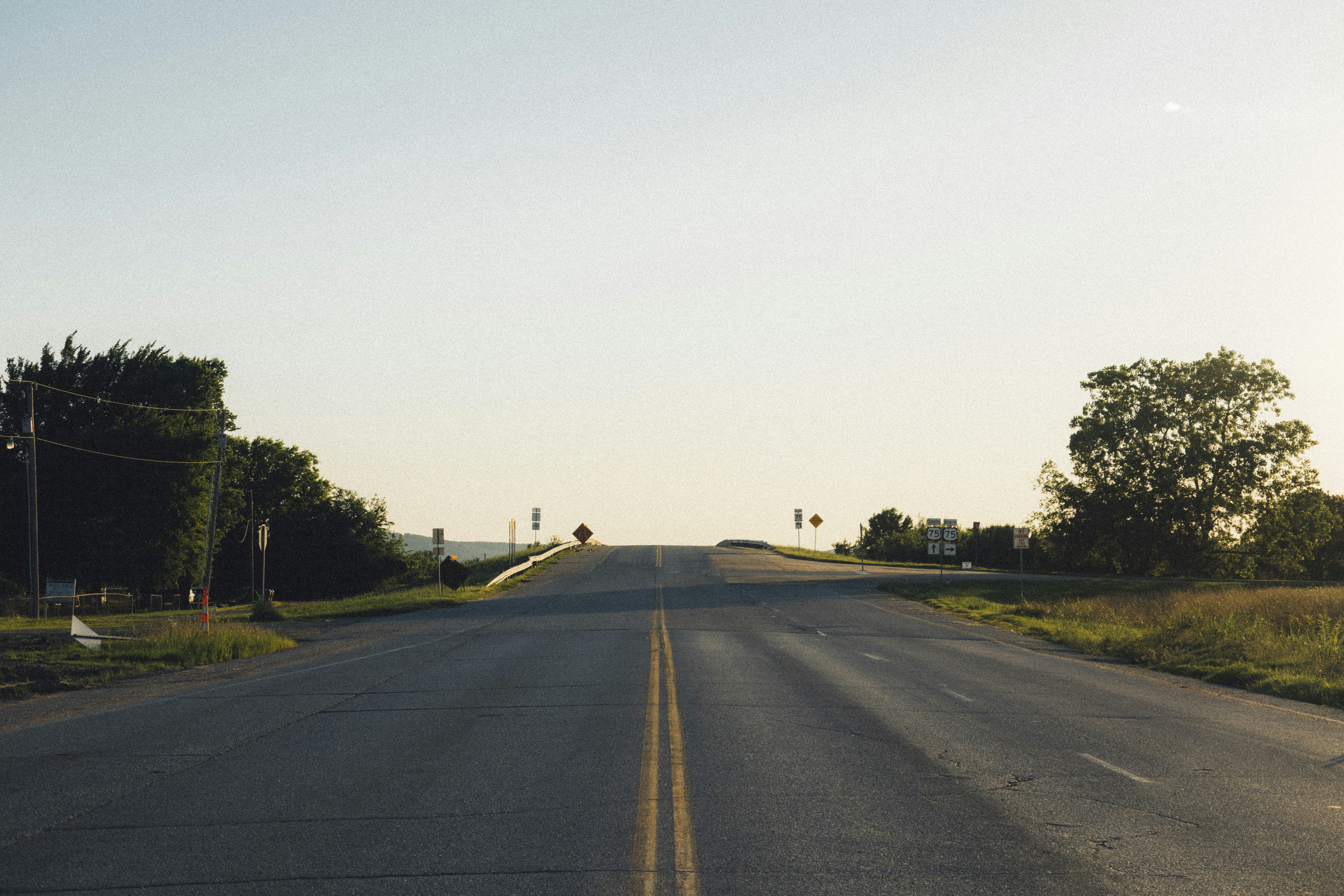 Empty road stretching toward the horizon, flanked by trees and utility poles under a soft evening light.