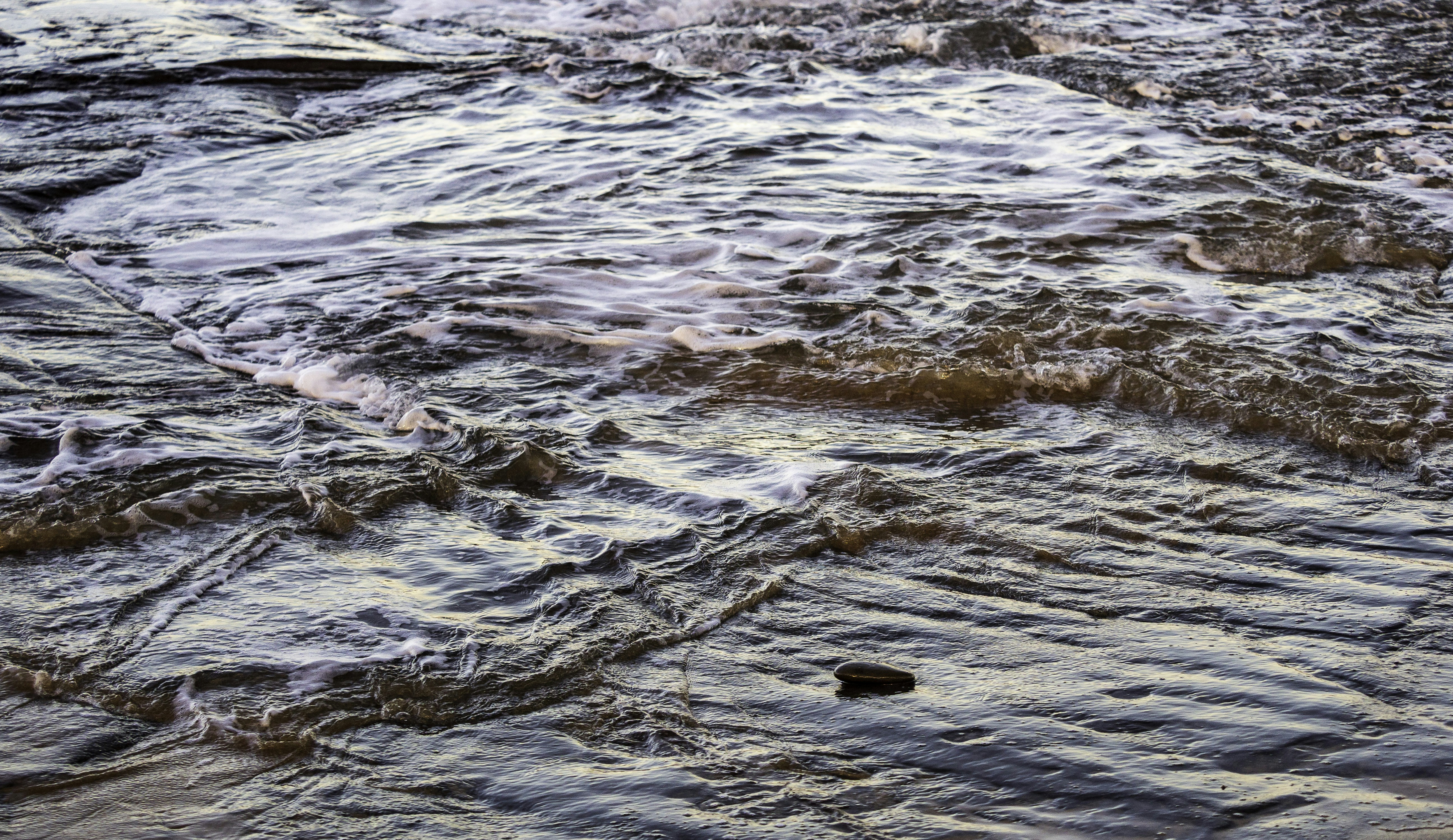 Gentle ripples and currents dance across a river's surface, revealing the interplay of light and water. A solitary stone rests below the flowing waves.