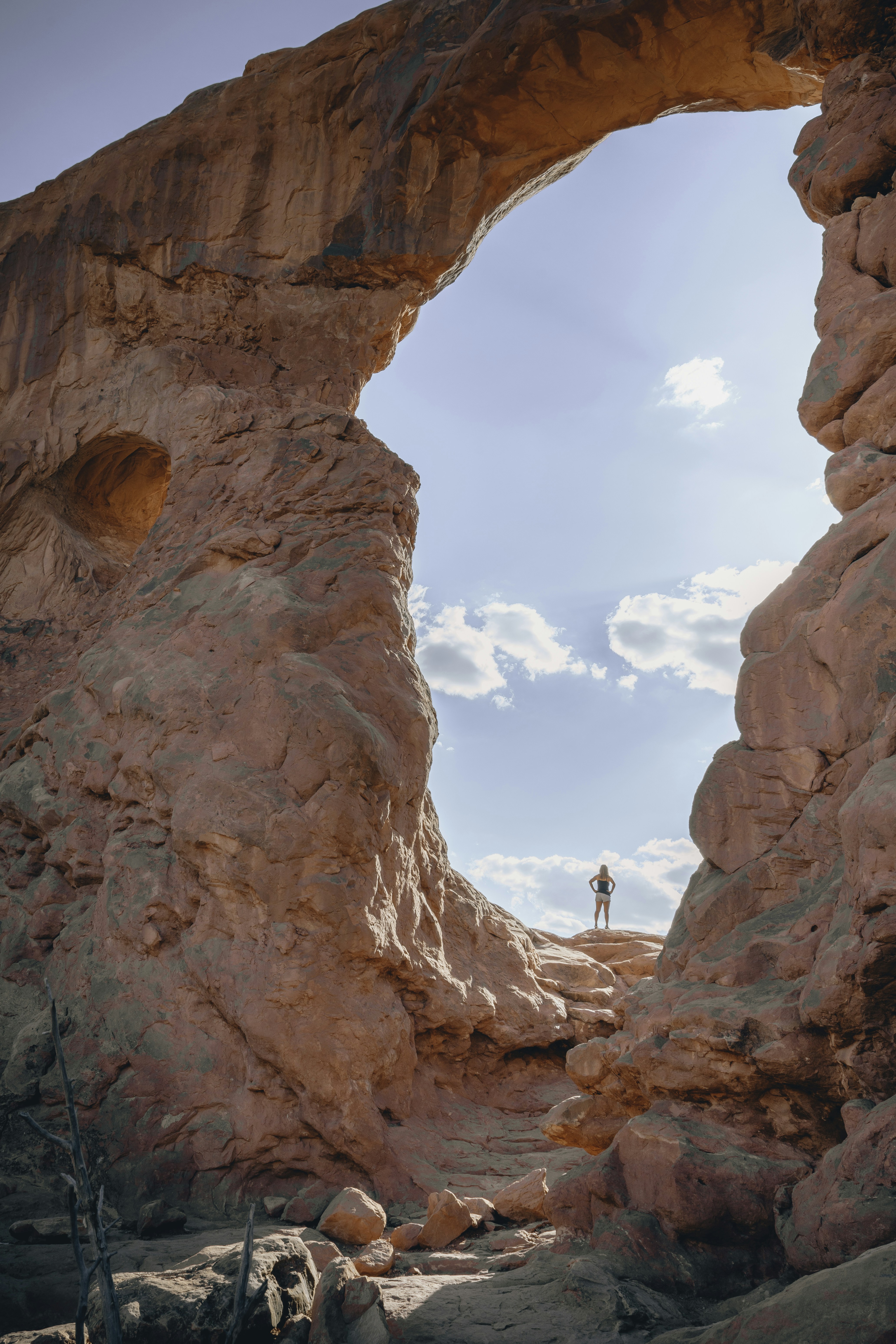 A lone figure stands at the base of a majestic rock arch, framed by rugged terrain and a bright blue sky dotted with clouds.