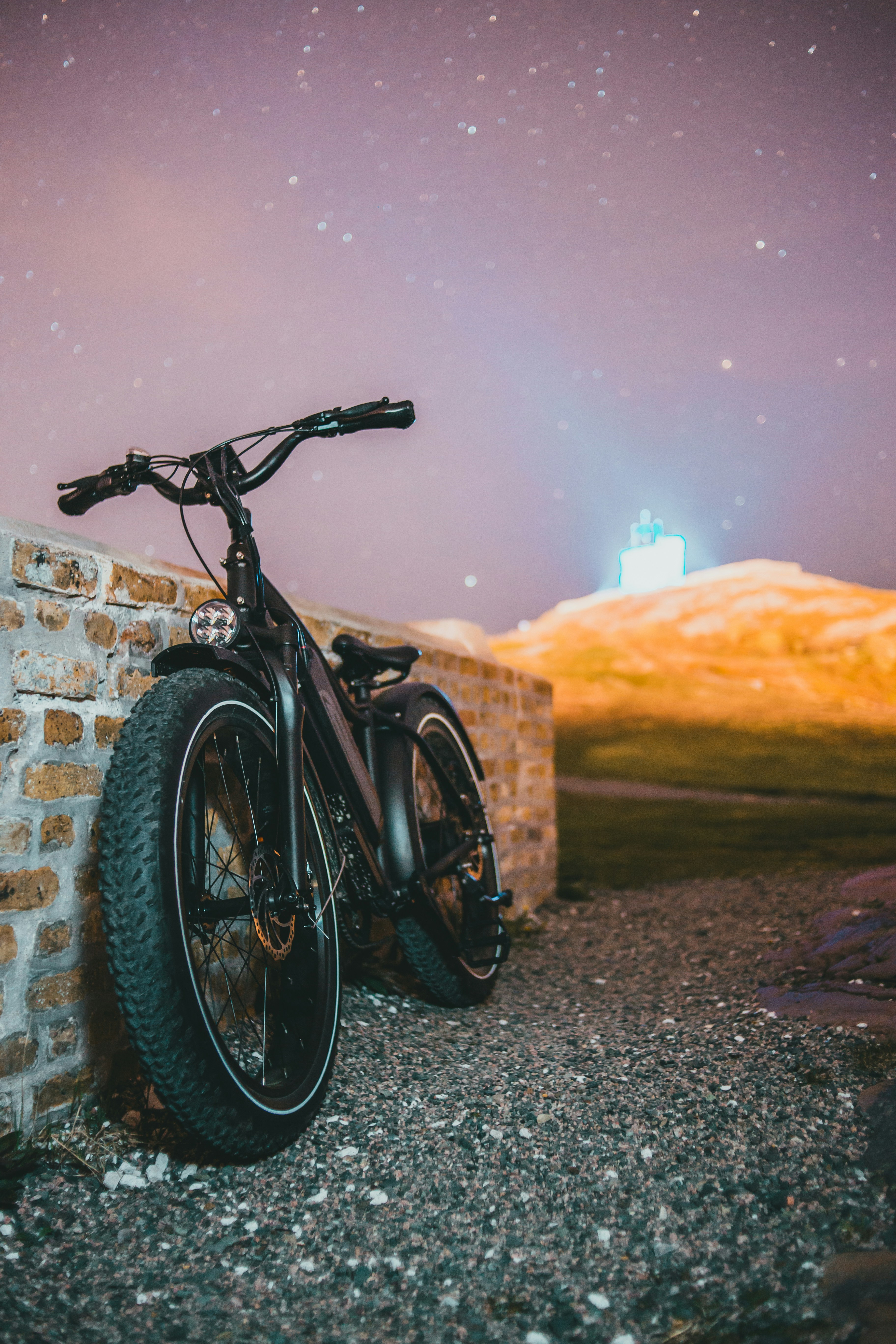 A sleek black bicycle rests against a textured stone wall, illuminated by a distant lighthouse under a starry sky.
