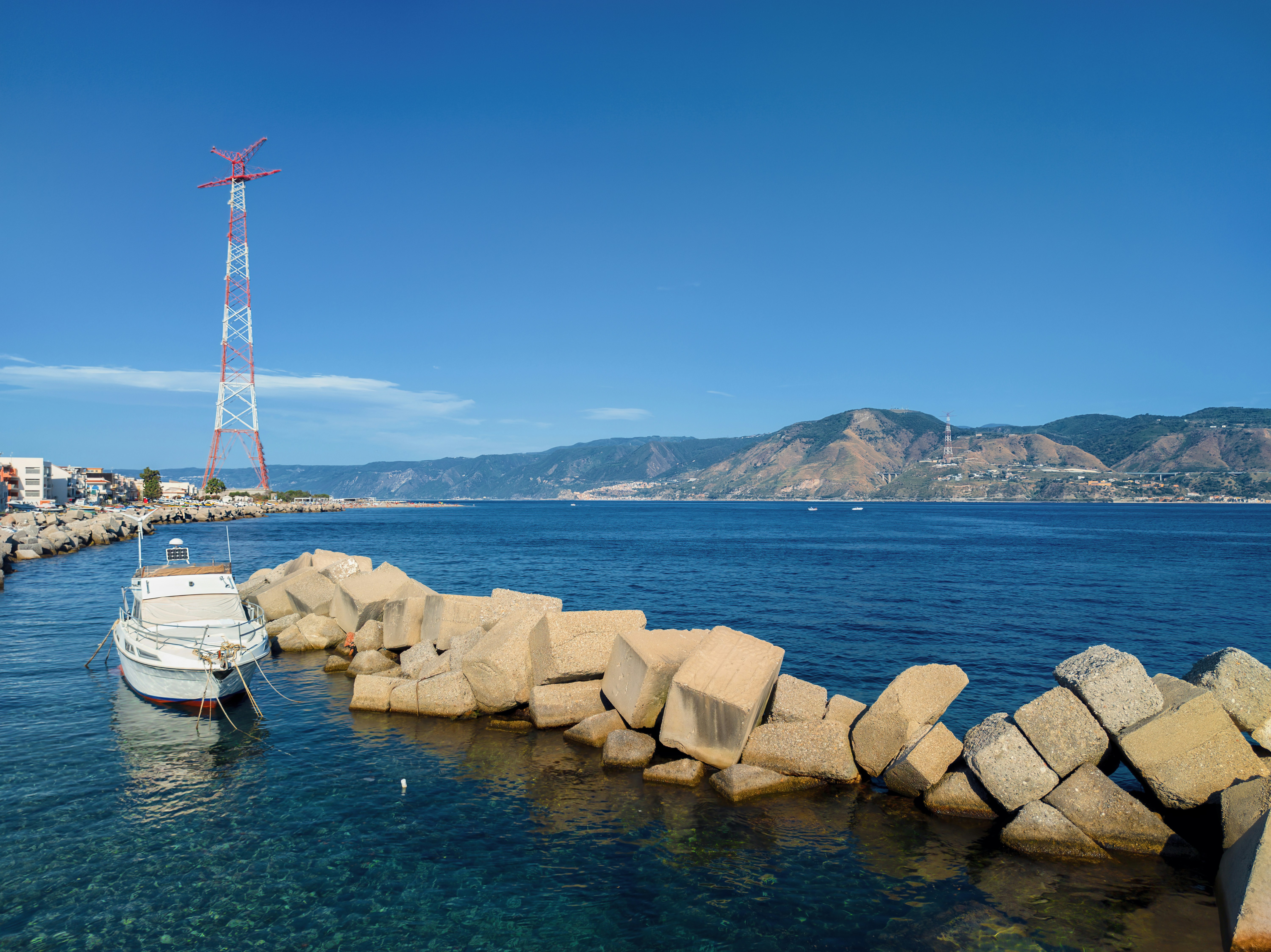 Fishing boat anchored beside a rocky breakwater with a distant mountainous backdrop and a towering radio mast. 