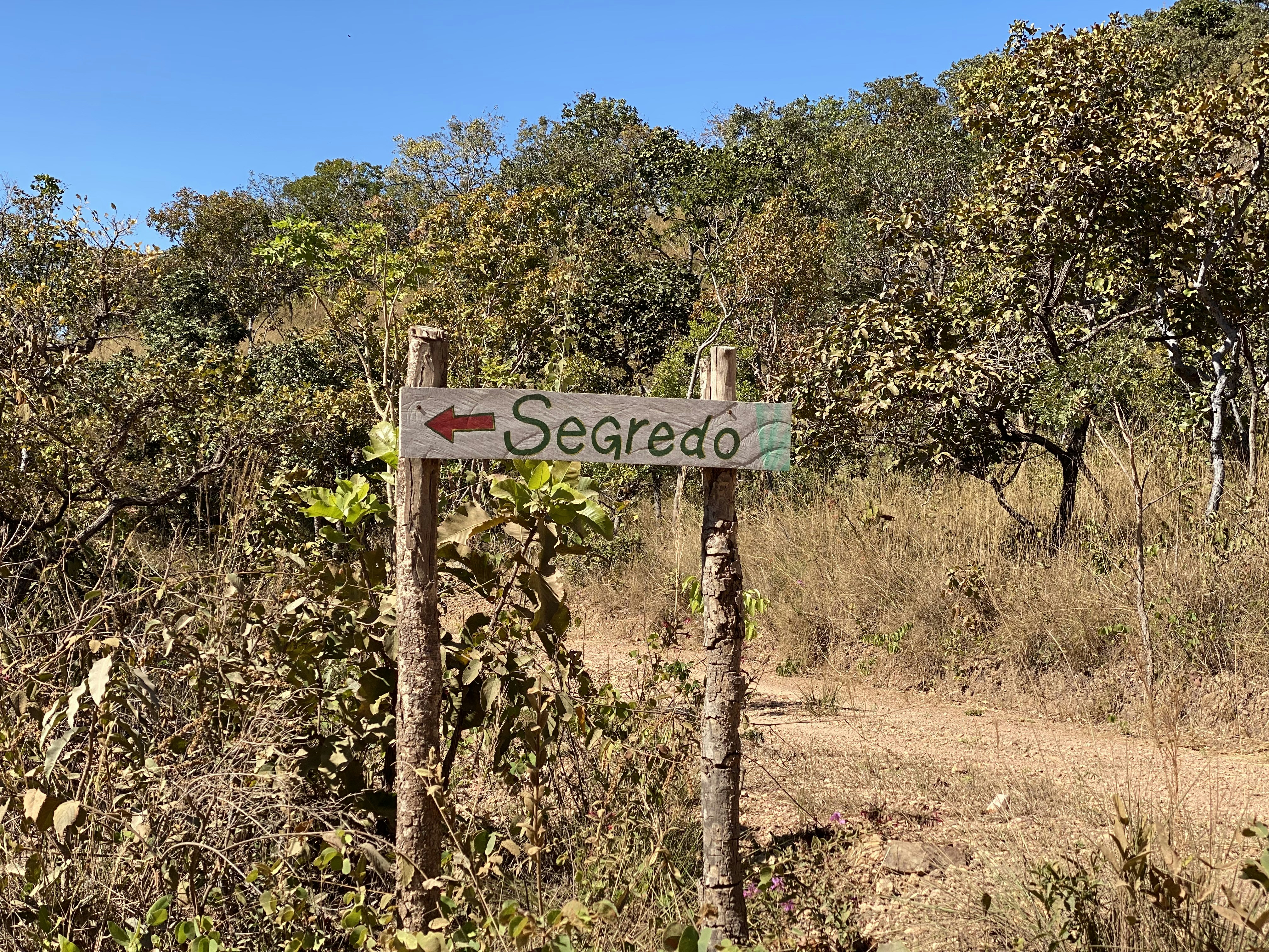 Wooden sign pointing towards 'Segredo' amidst a dry, bushy landscape under a clear blue sky.