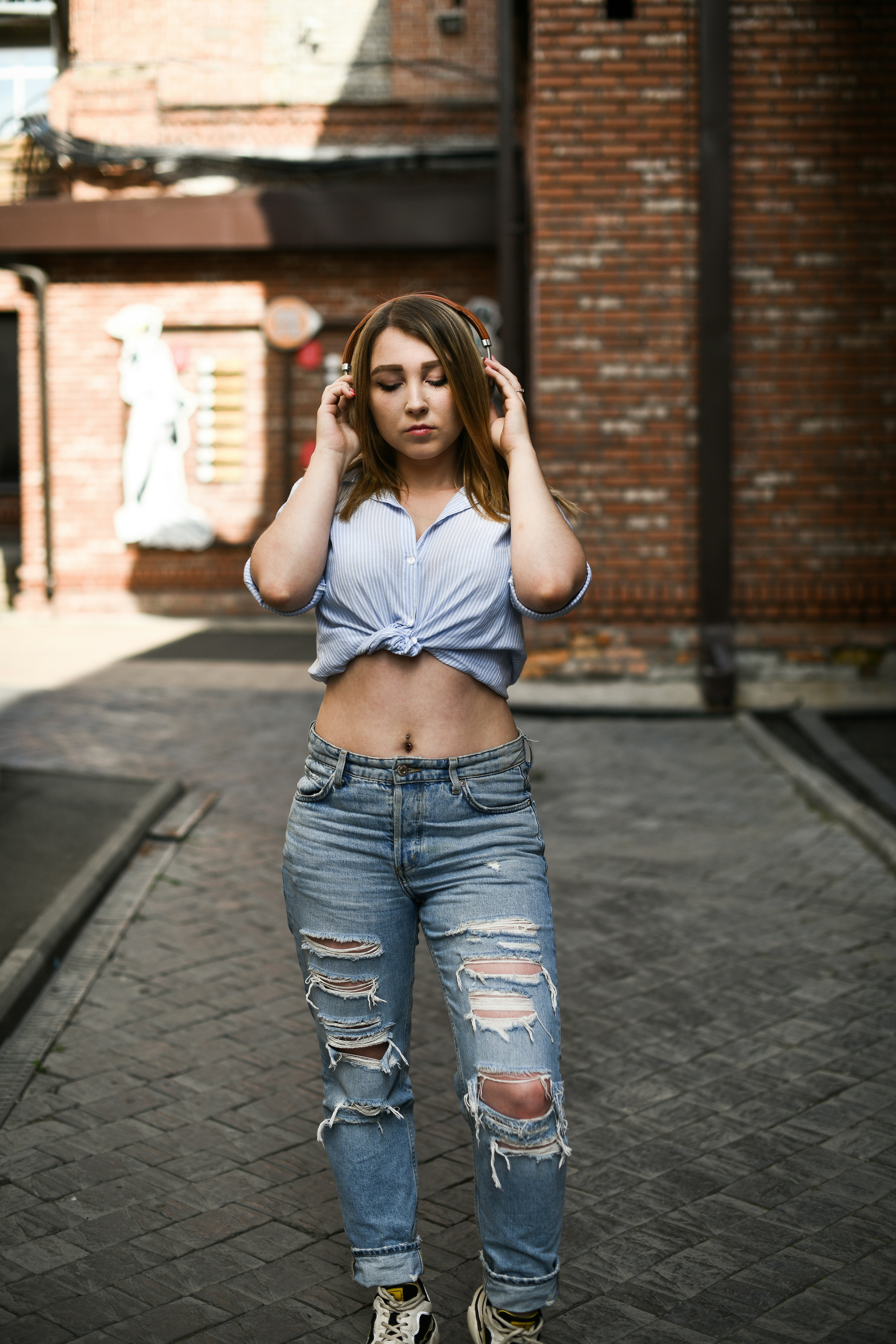 Young woman in a striped shirt and ripped jeans stands in an urban setting, adjusting her headphones. The backdrop features rustic brick architecture.