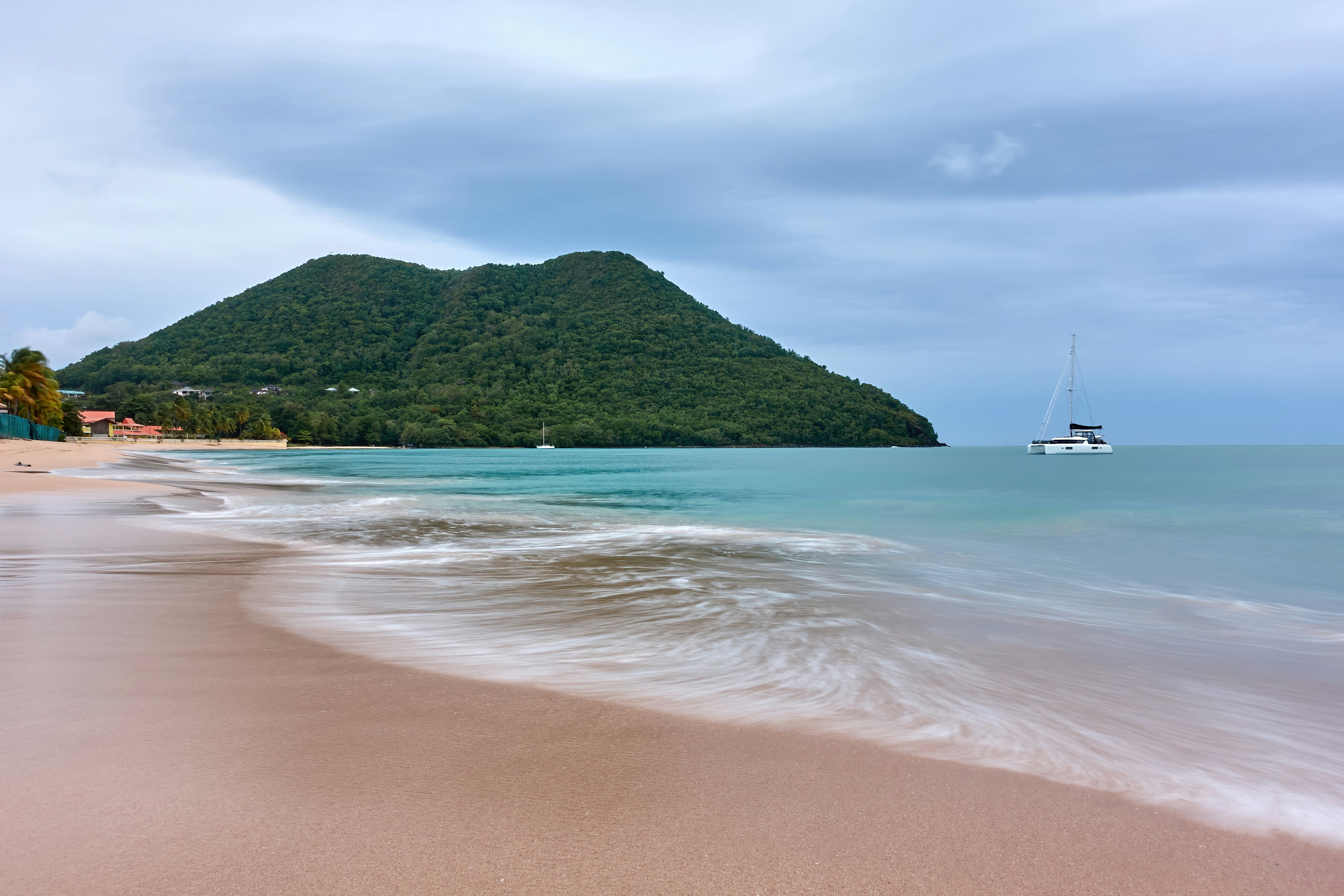 Gentle waves lap against a sandy beach under a cloudy sky, with a lush green hill and anchored sailboat in the distance.