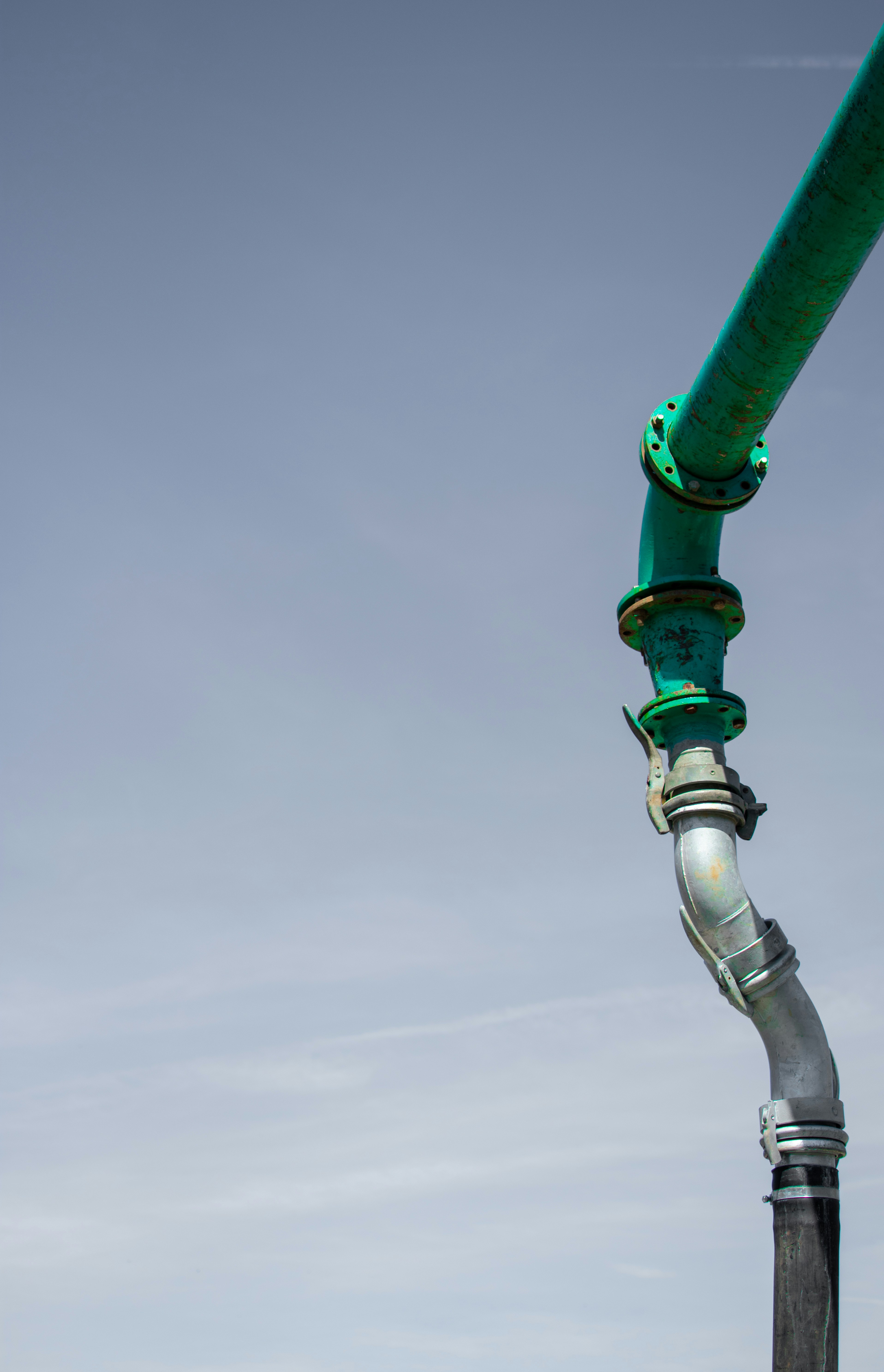 Industrial teal pipe with flanges arcs against a pale blue sky, emphasizing weathered metal and joints.