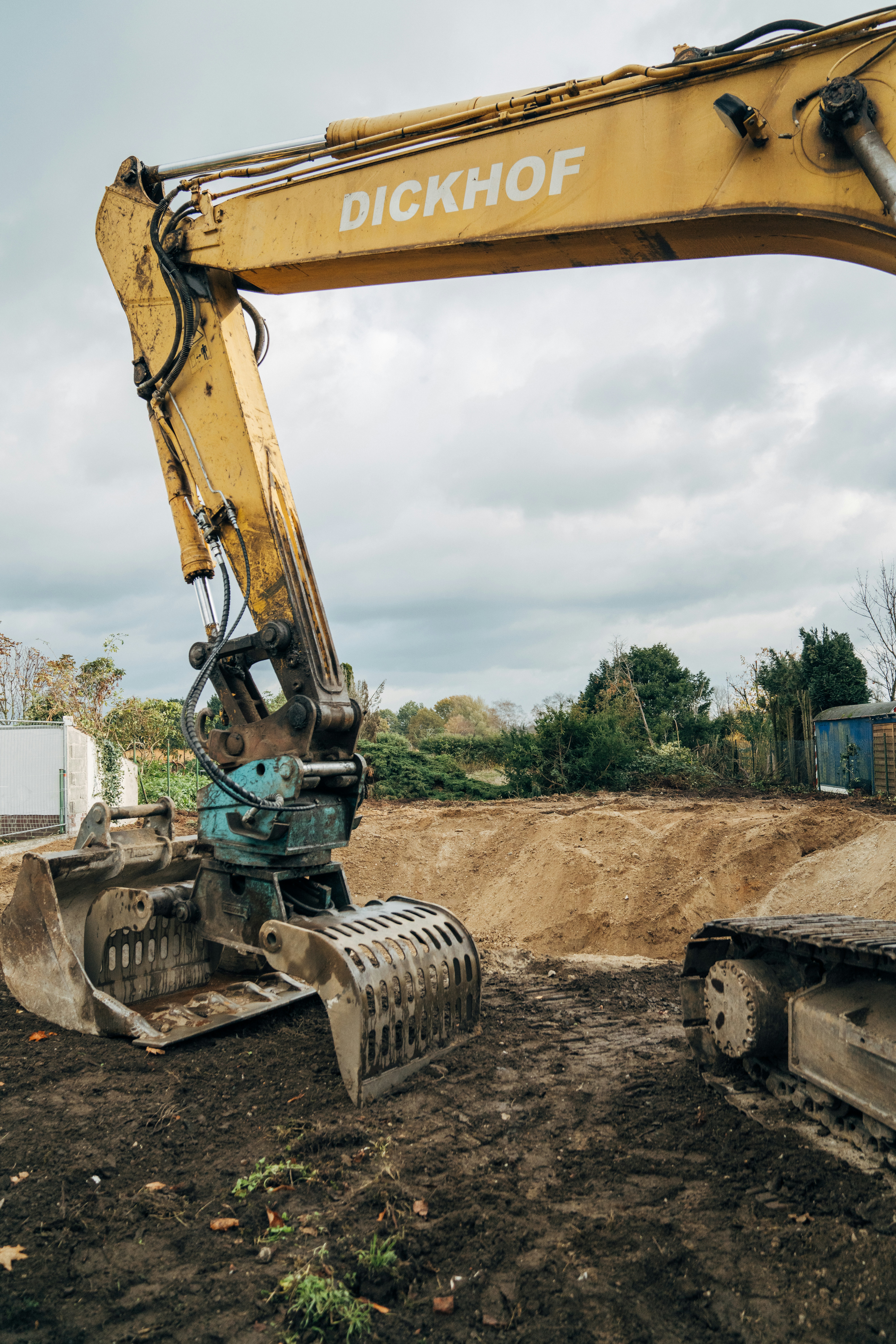 Excavator with a prominent yellow arm and gripping claws poised for action at a construction site, surrounded by earth and greenery.