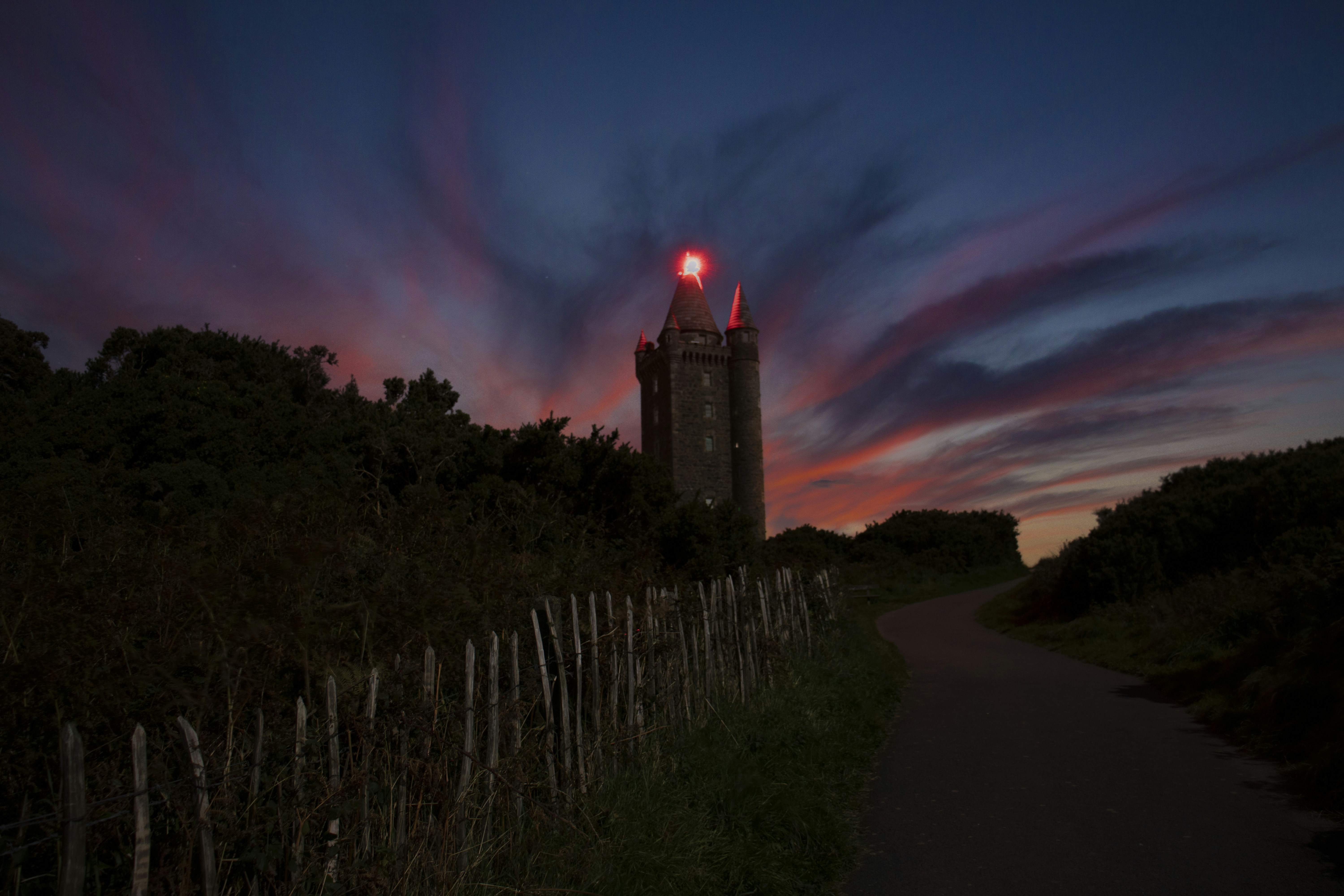 A historic tower stands silhouetted against a vibrant twilight sky, with wispy clouds painted in shades of pink and blue. The tower's beacon glows brightly, guiding the way for evening wanderers.