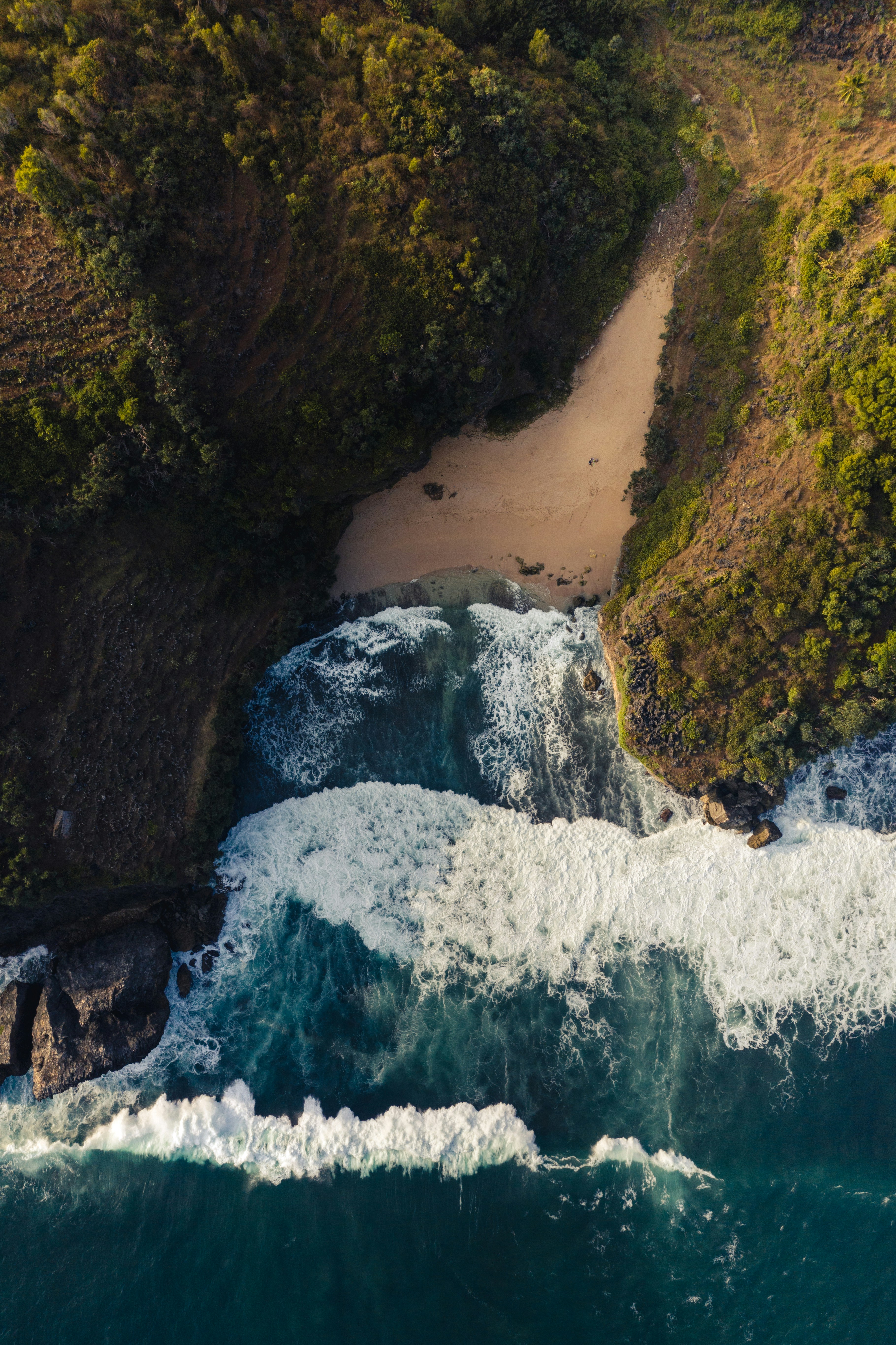 Aerial view of a secluded beach nestled between lush green hills and crashing waves, showcasing the serene beauty of a hidden cove.