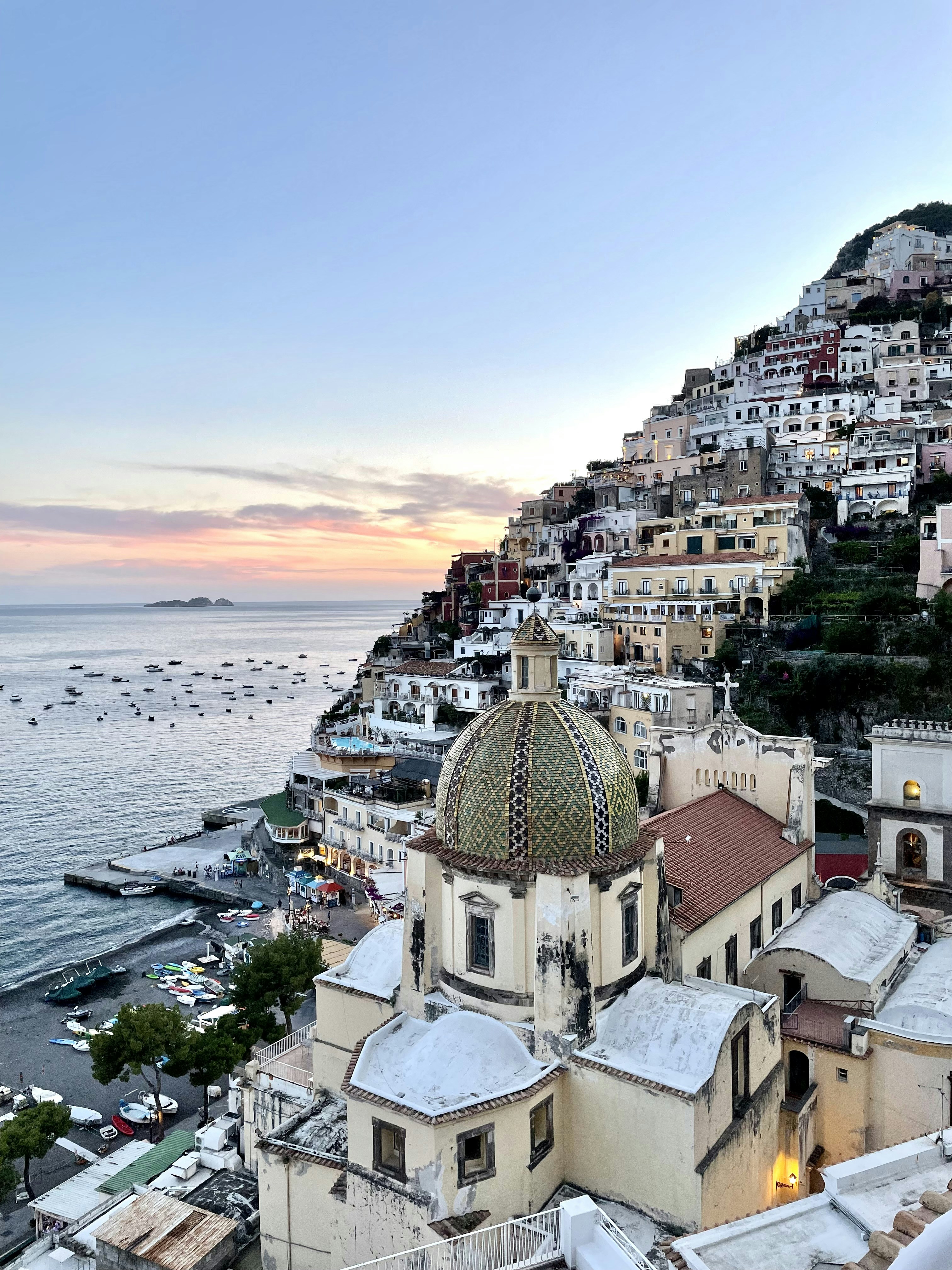 Vibrant coastal village of Positano at sunset, showcasing terraced buildings and a distinctive domed church. The serene sea reflects the evening hues.