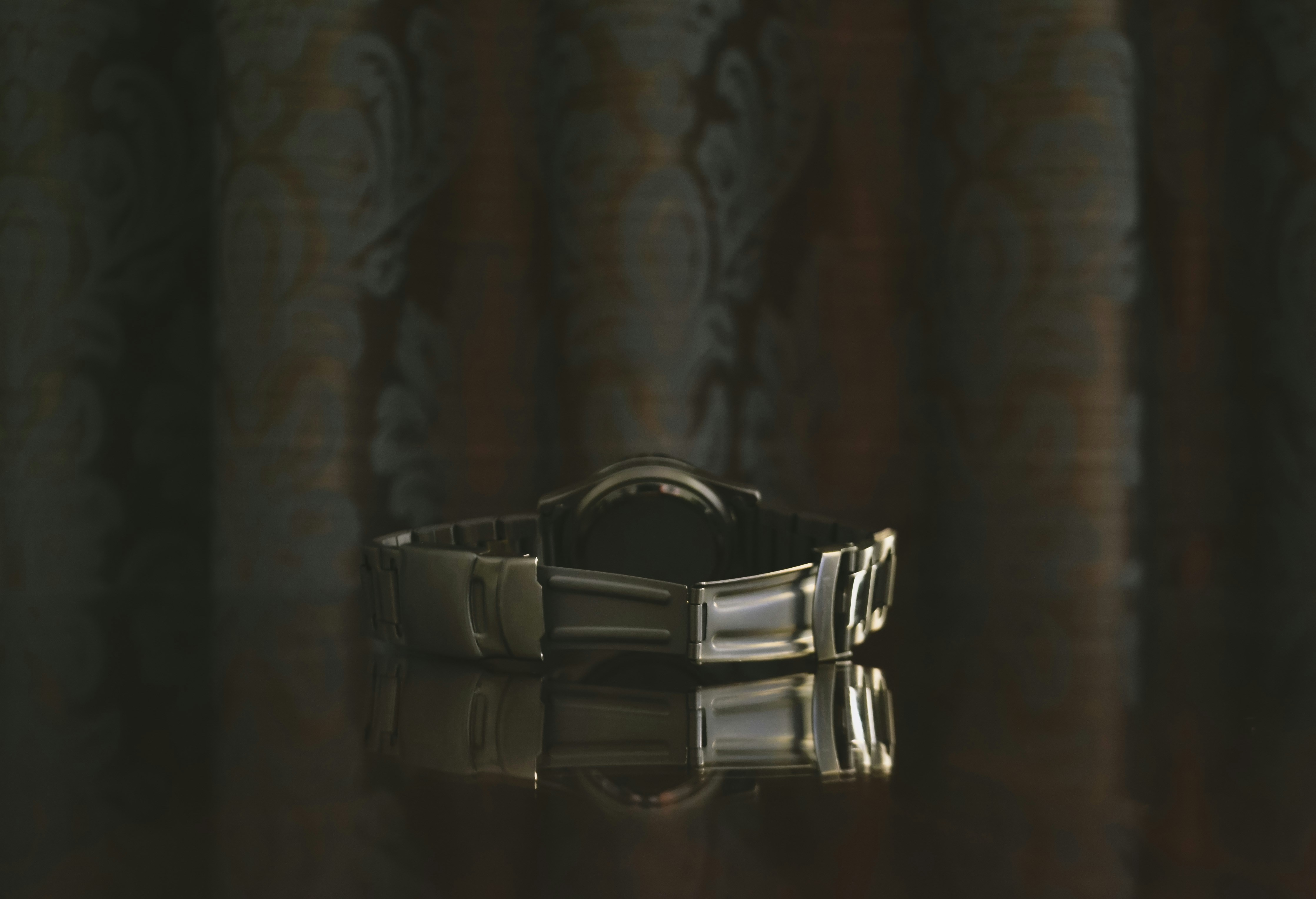 Close-up of a metallic watch resting on a reflective surface, partially obscured by patterned curtains in the background.