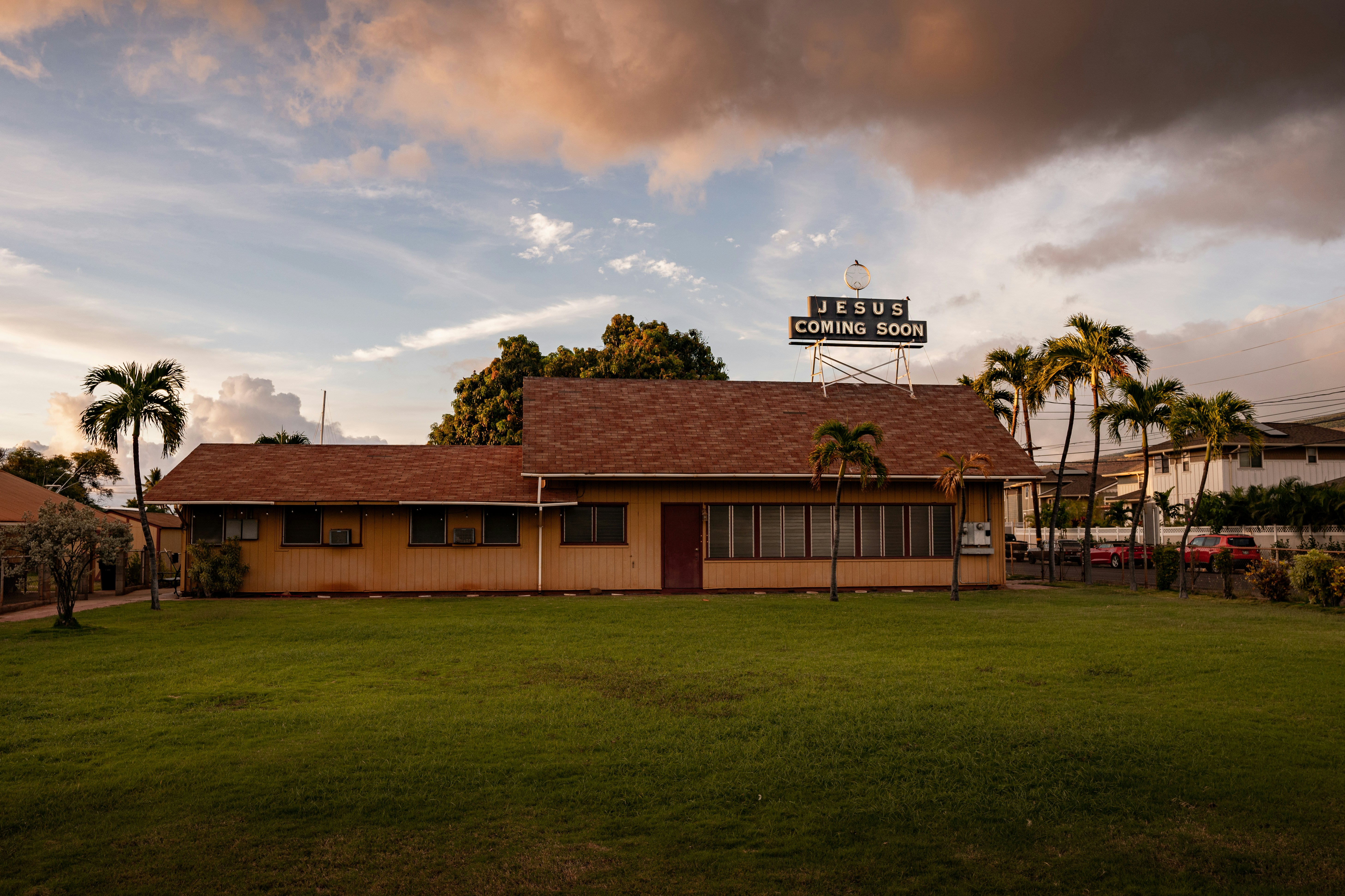 A quaint building with a sign reading 'Jesus Coming Soon' stands amidst lush green grass and palm trees under a dramatic sky.