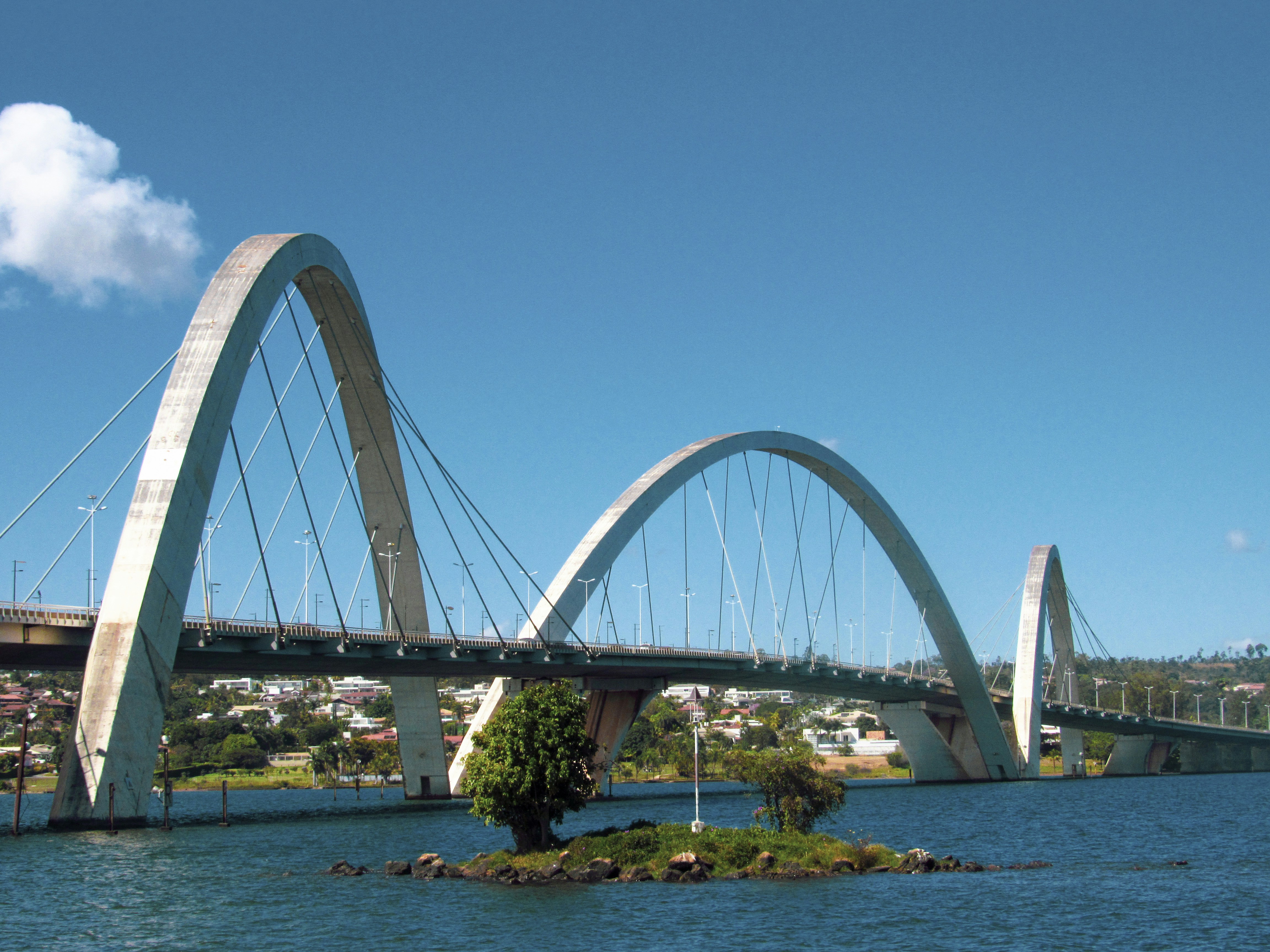 A striking modern bridge with sweeping arches spans a tranquil body of water, surrounded by lush greenery and a clear blue sky.