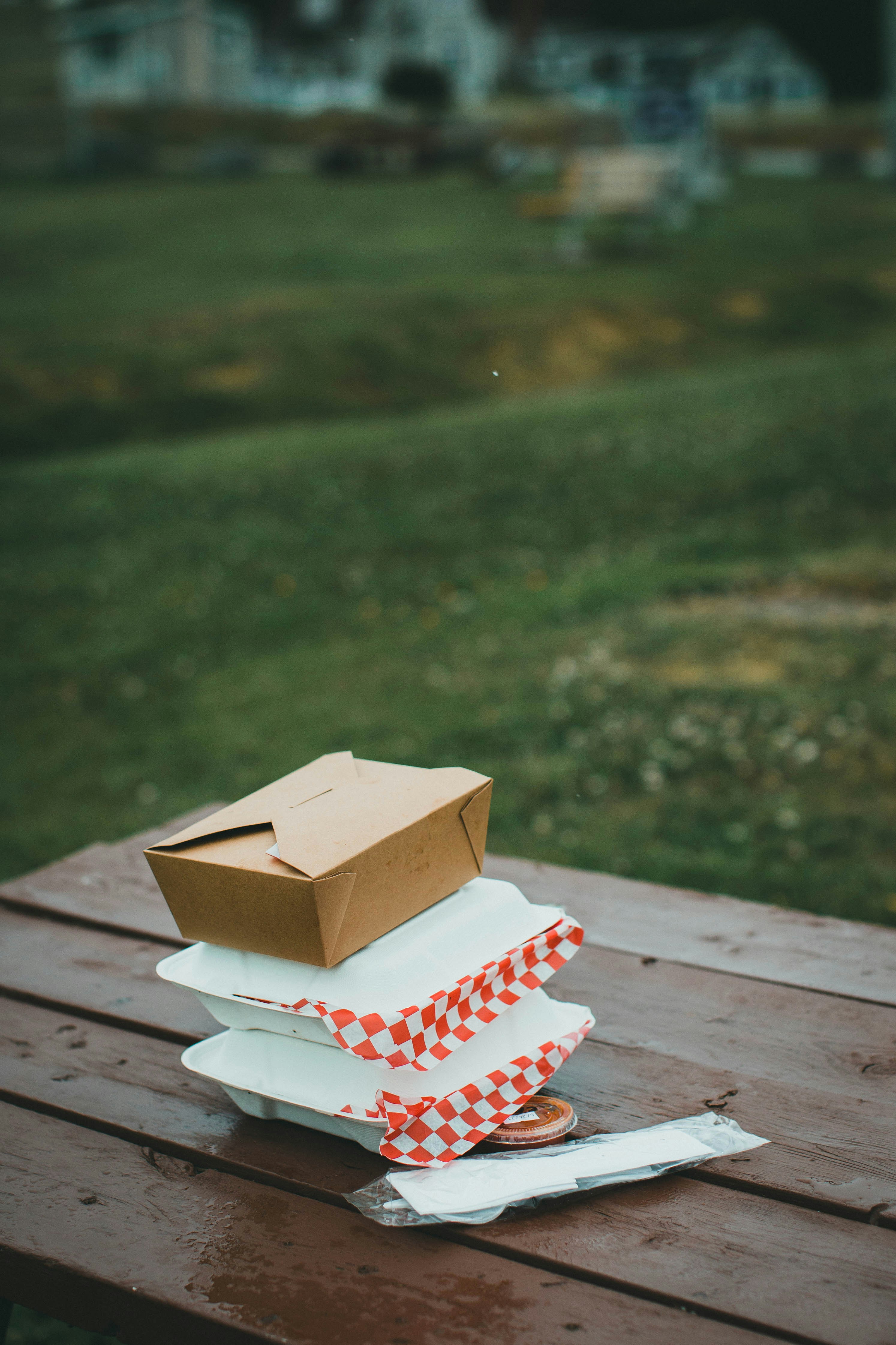 Stack of takeout containers on a wooden table, with a blurred background of a grassy area. The scene captures the essence of casual dining.