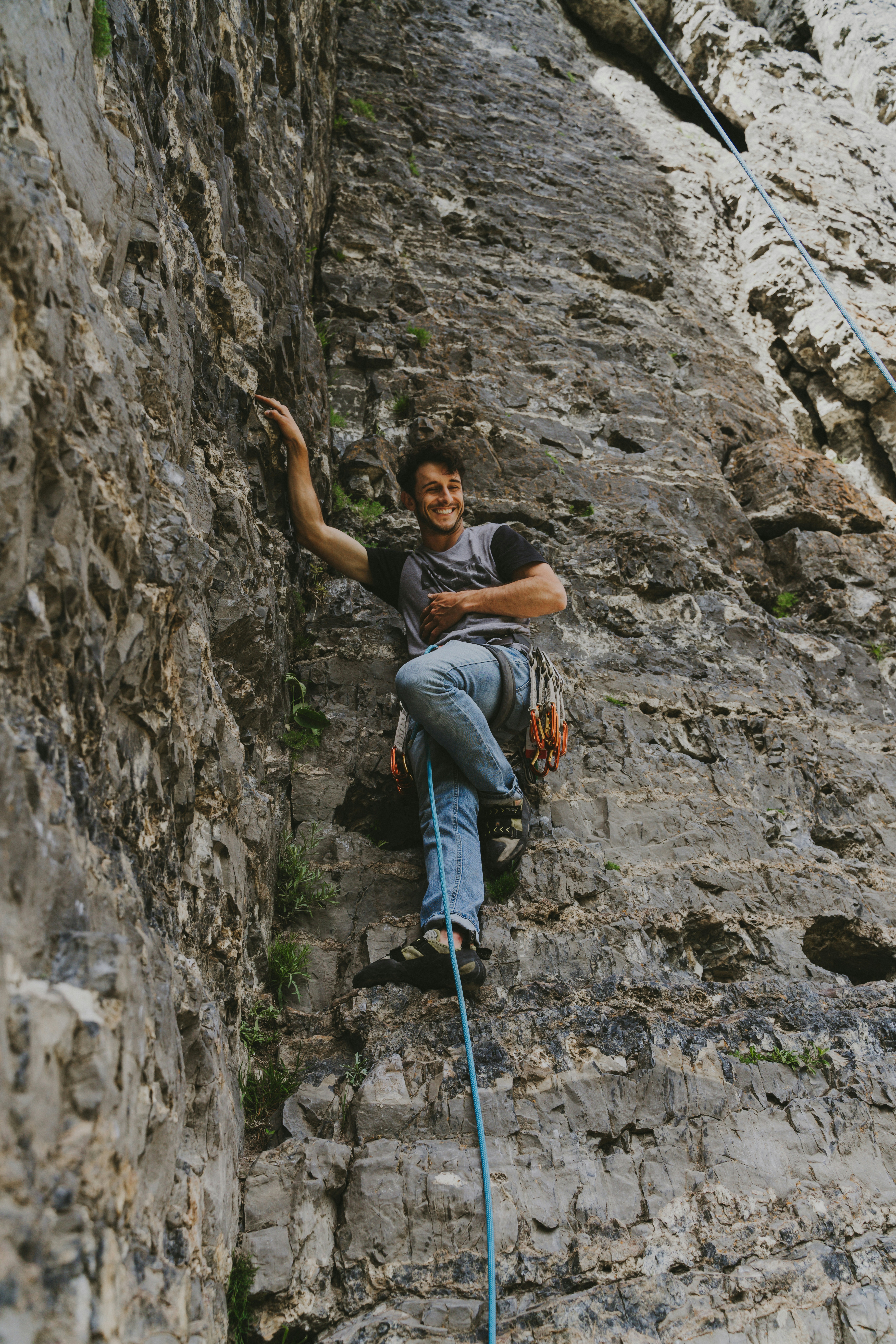 Climber resting against a rocky wall, smiling while secured by climbing gear and rope. The rugged texture of the stone contrasts with the climber's relaxed posture.