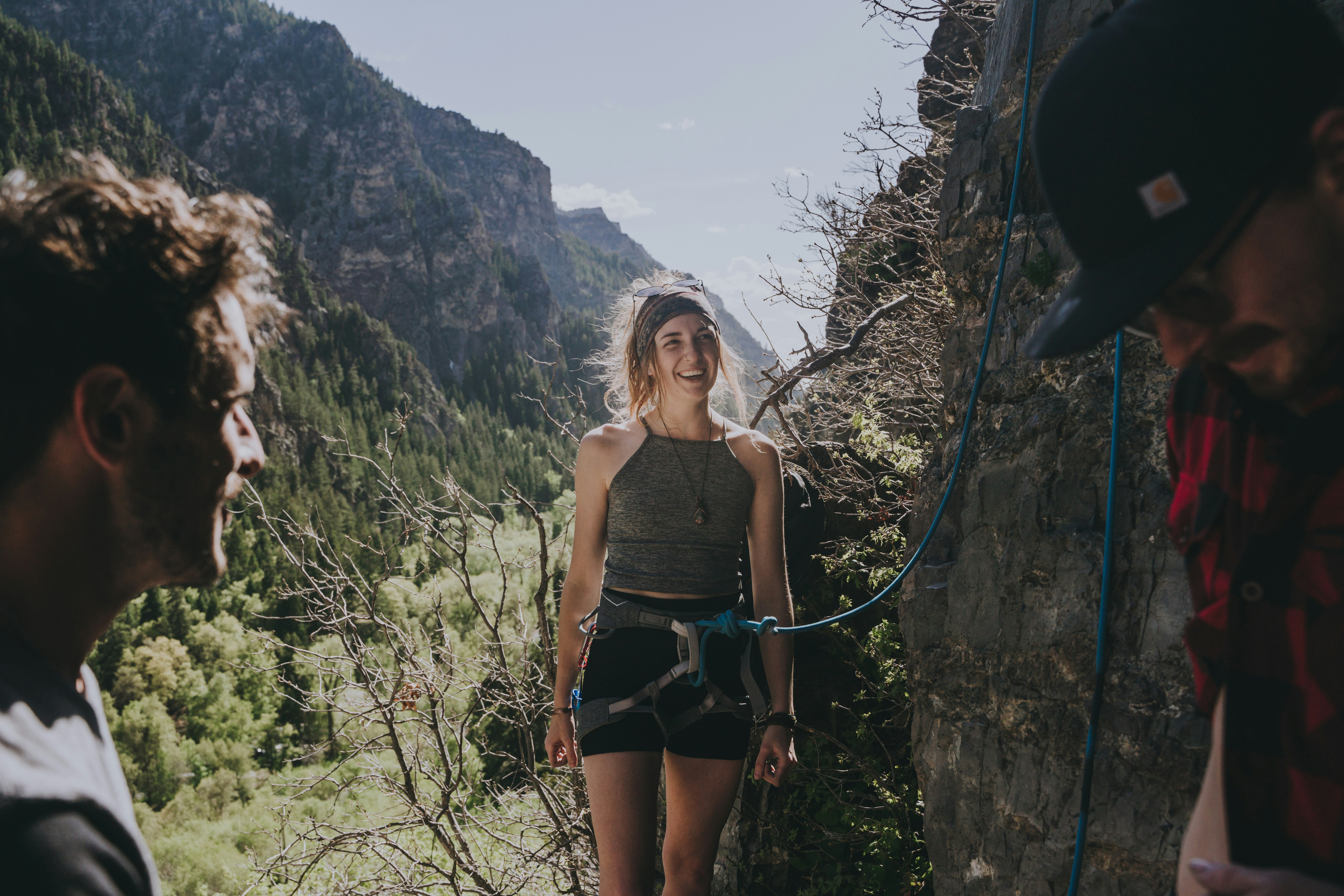 Climber smiling while secured to a harness on a rocky cliff, surrounded by lush green mountains under a clear blue sky.