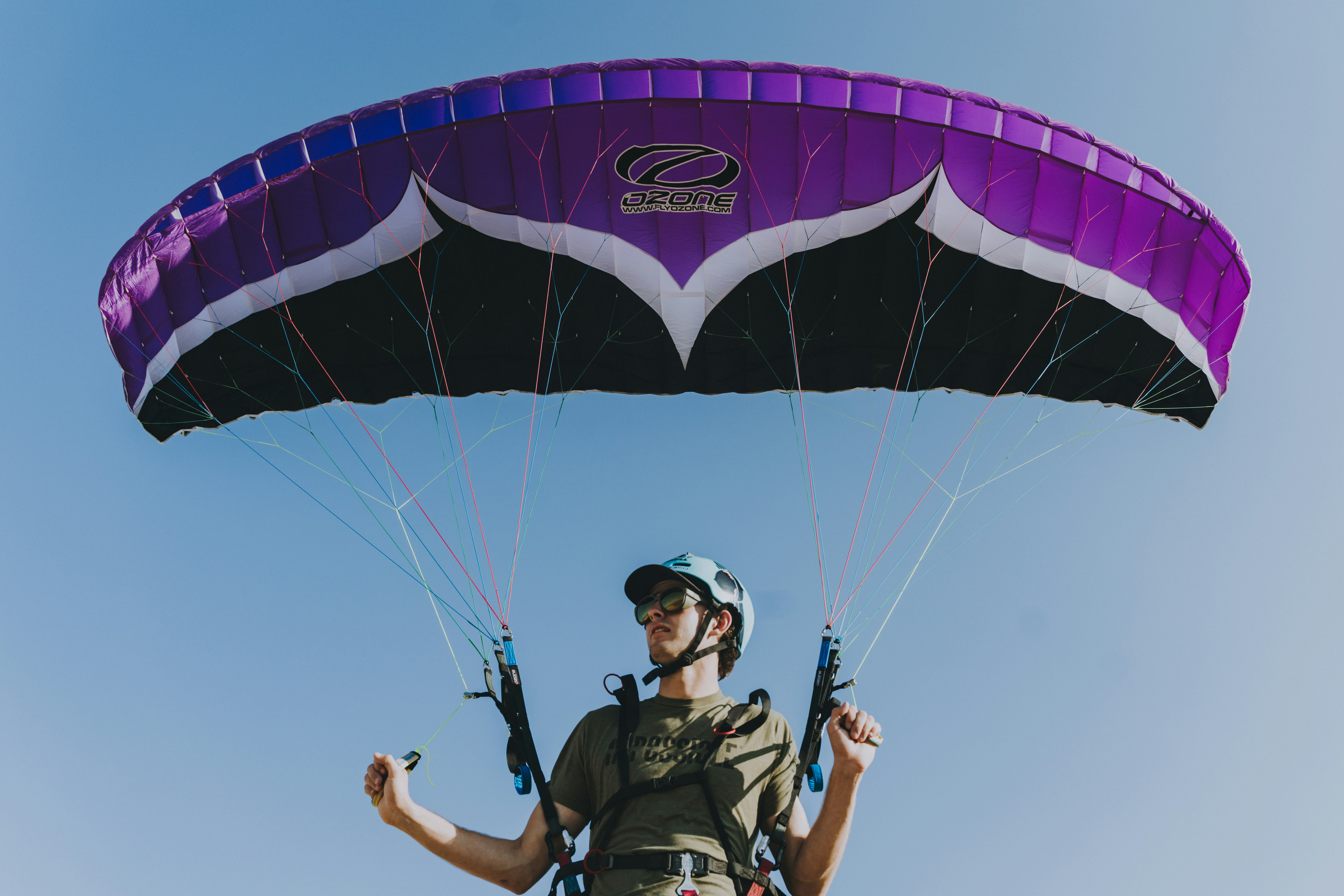 Paraglider technician ascends under a vivid purple wing, harness and helmet visible against clear blue sky. A mid-air moment emphasizing control and gear preparation.