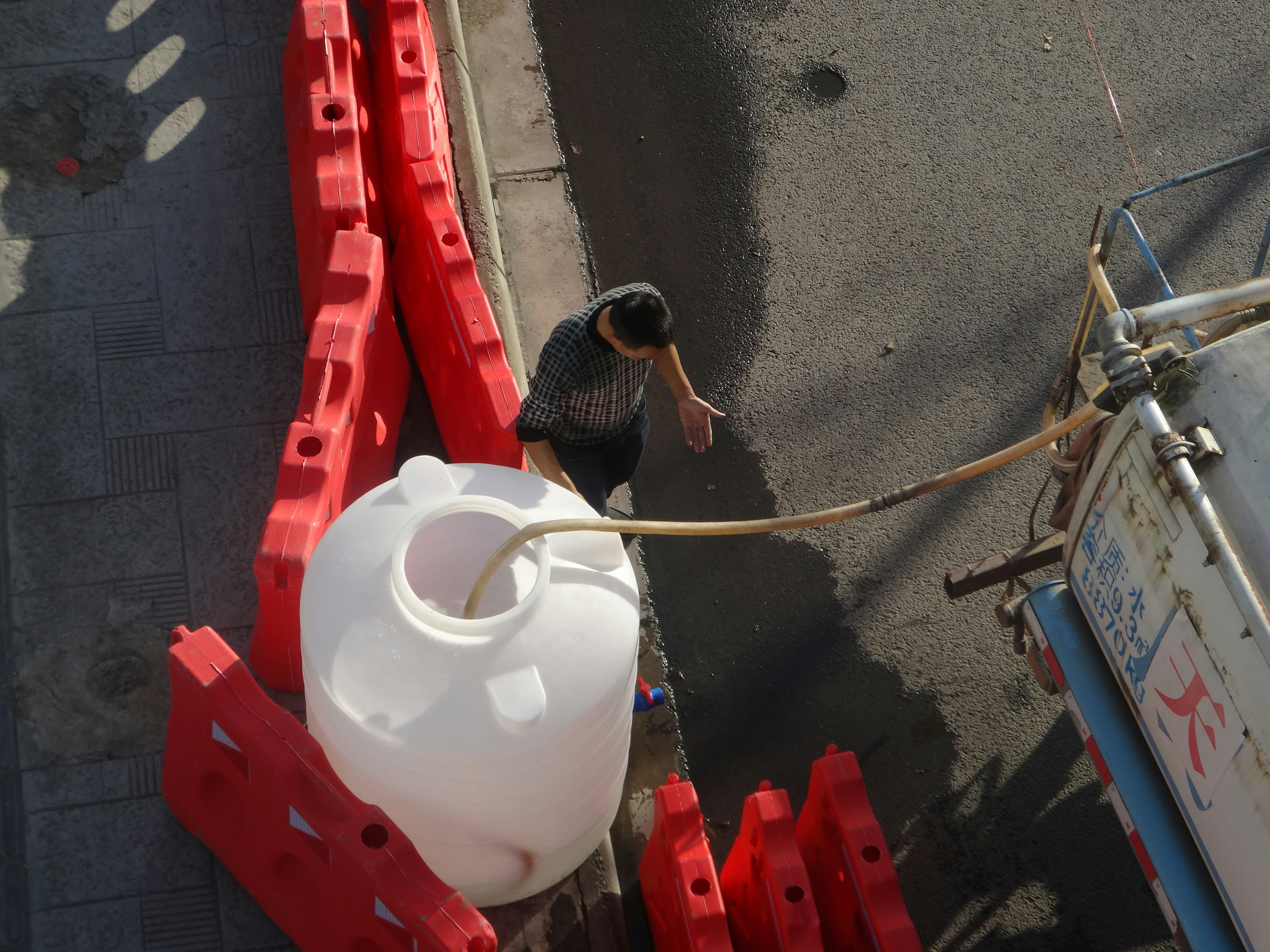 Worker refilling a large white container from a truck, surrounded by red barriers on a street.