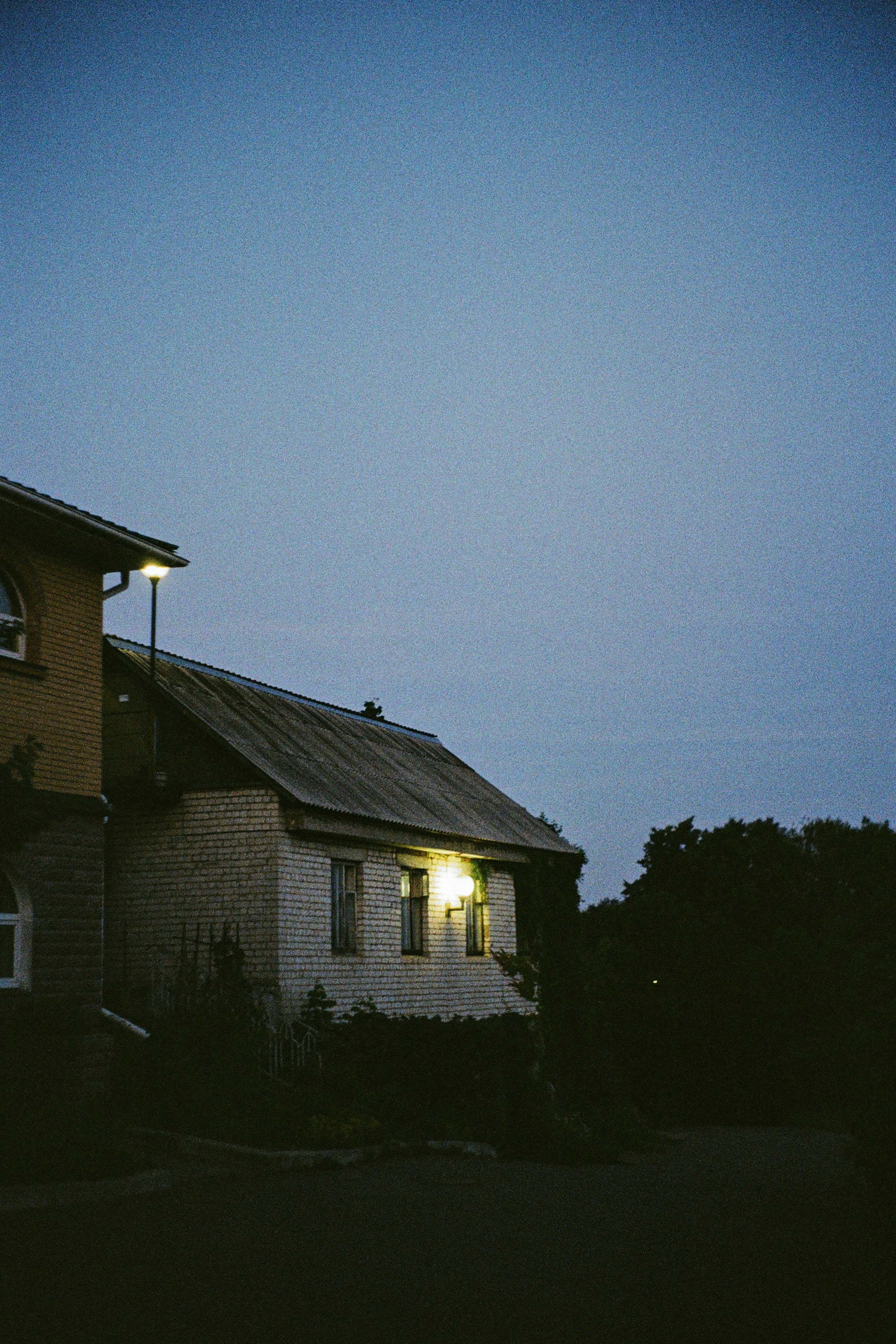 A dimly lit house stands under a twilight sky, with a single light illuminating its side. The tranquil scene captures the essence of evening calm.