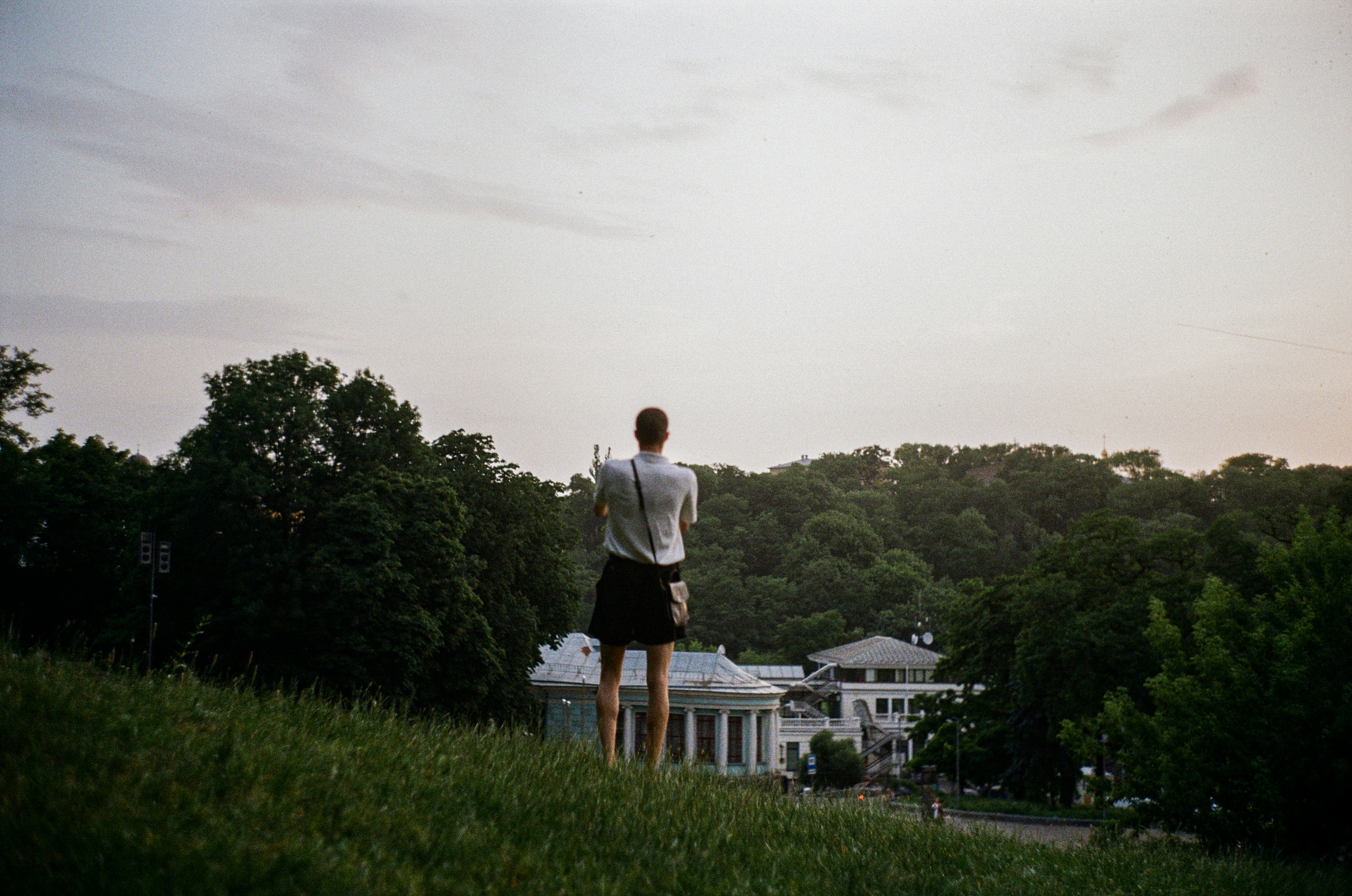 Hooded figure stands on a grassy hill facing a distant stately building surrounded by trees, captured in soft evening light. A calm, expansive scene with a subtle human presence highlighting solitude.