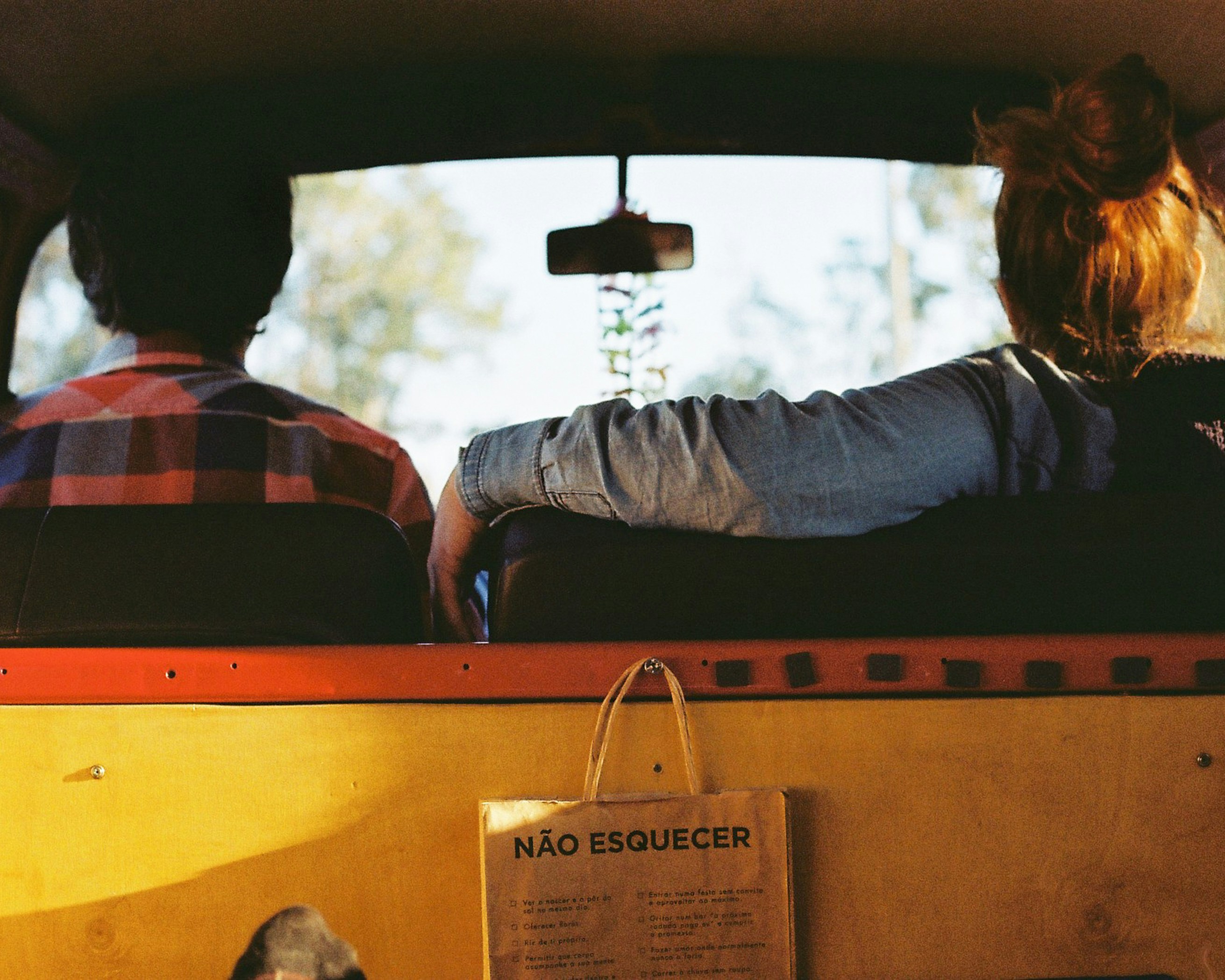 Two individuals seated in a vintage vehicle, enjoying the view ahead. A bag with the phrase 'NÃO ESQUECER' hangs from the back seat.