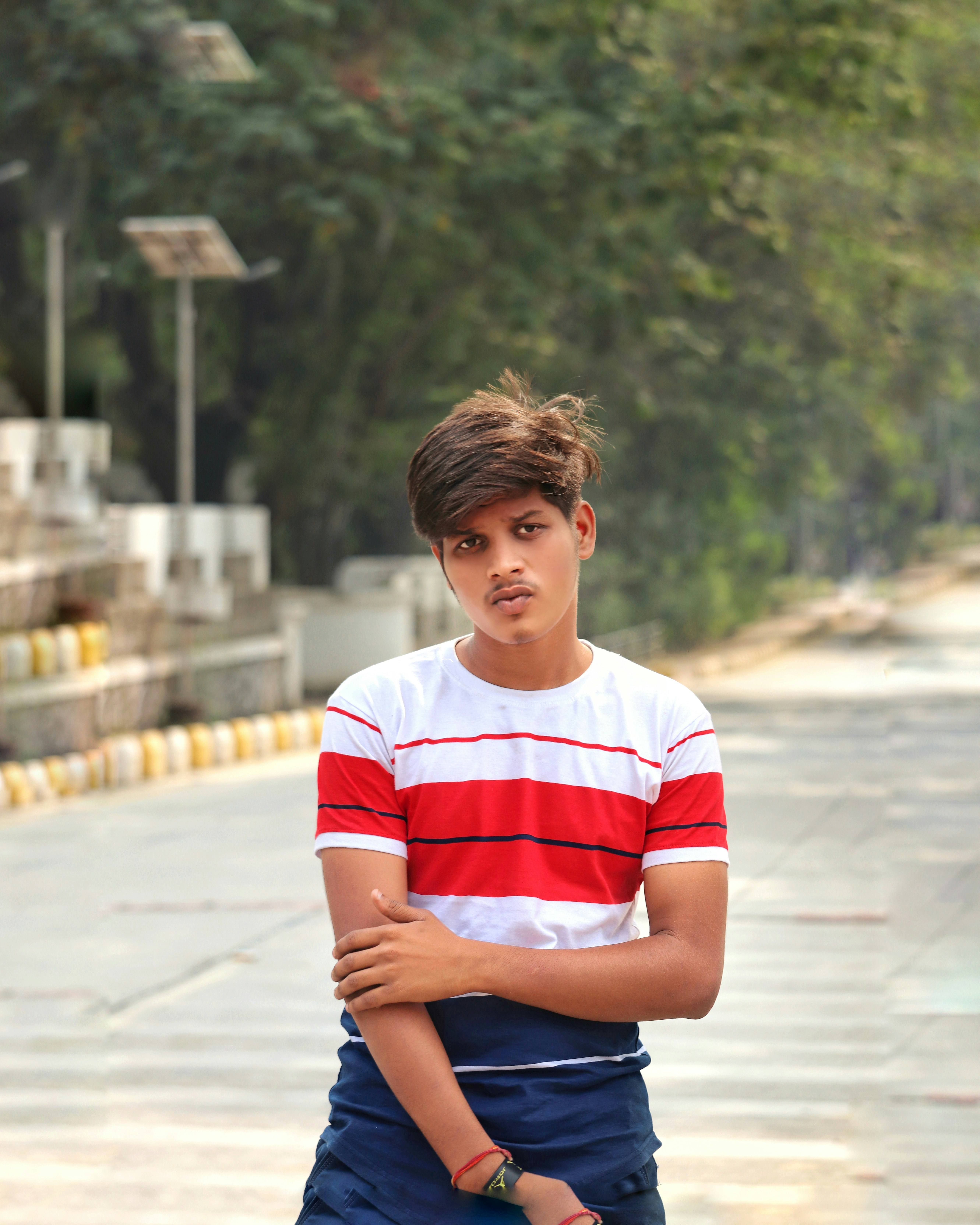 Young man in a red and white striped shirt stands thoughtfully on a quiet street, surrounded by greenery and soft natural light.
