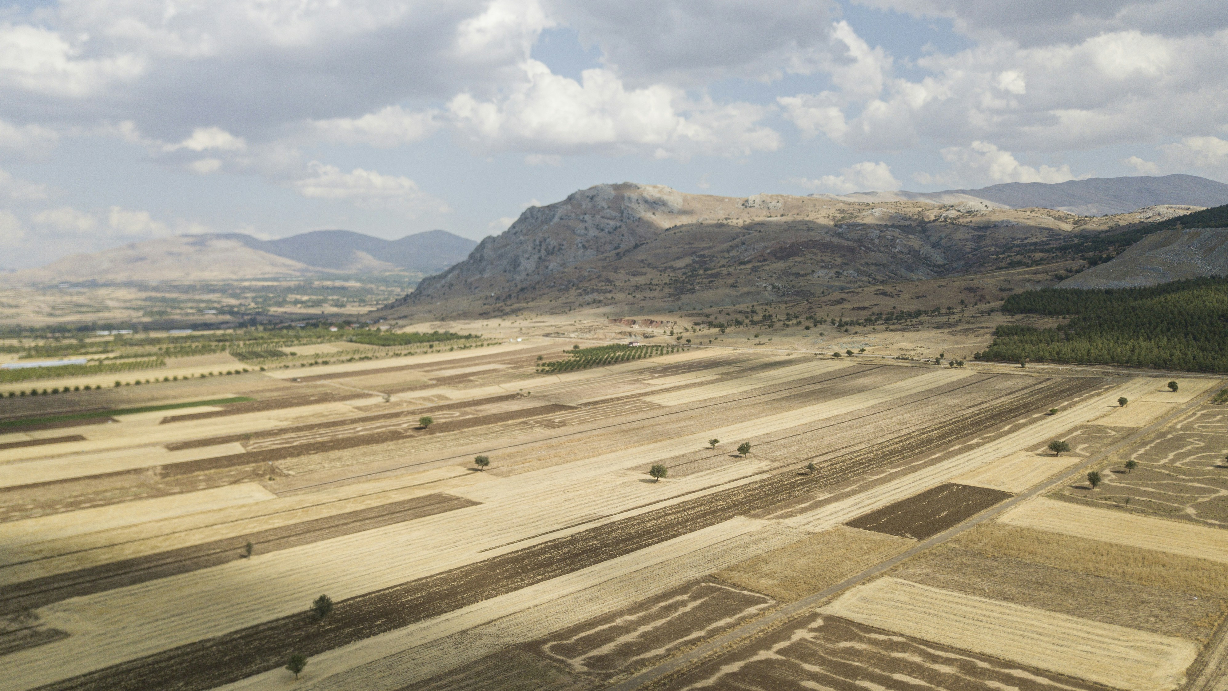 Expansive agricultural fields spread out beneath a rugged mountain under a cloud-speckled sky.