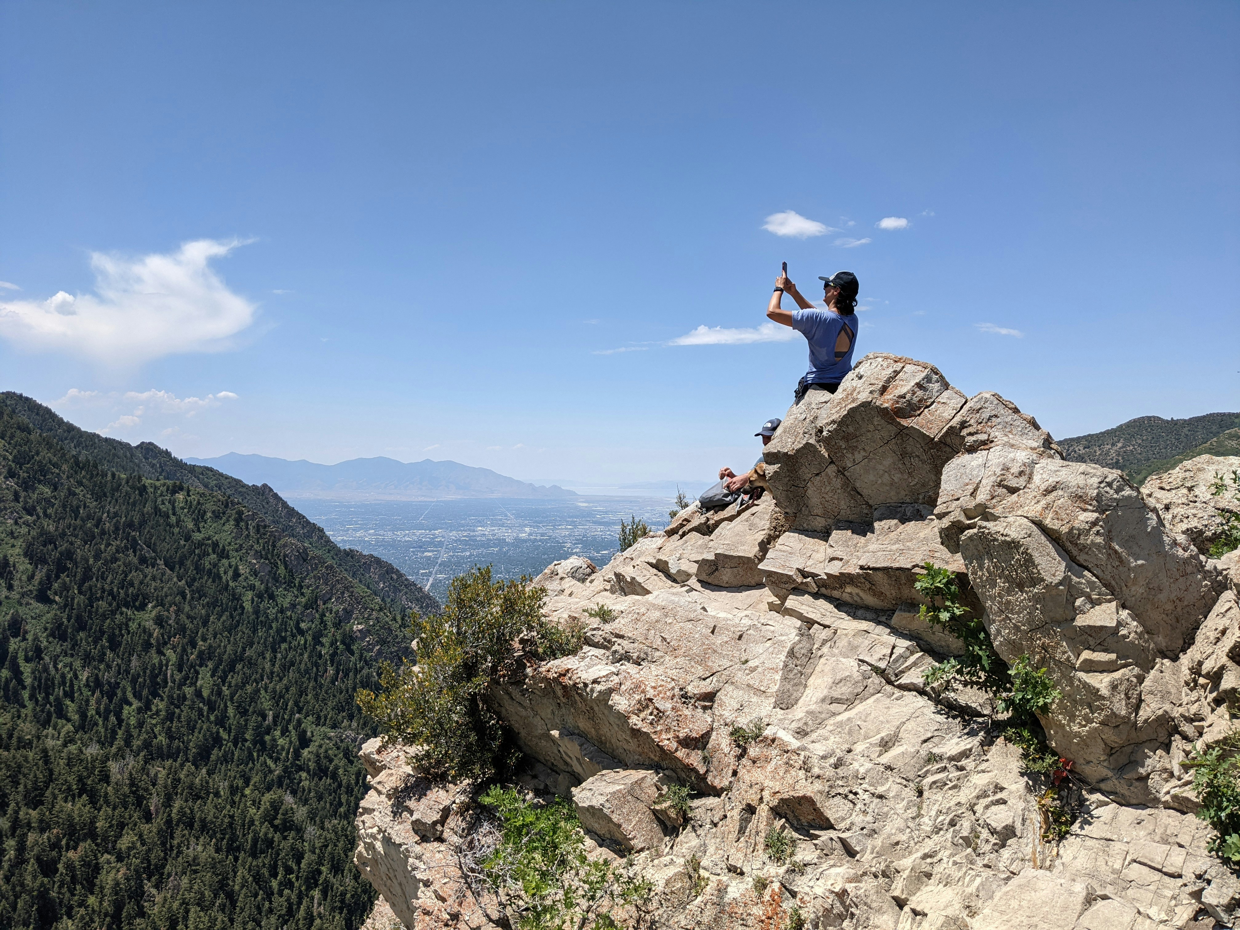 Individual perched on a rocky outcrop, capturing a panoramic view of the valley below, surrounded by lush greenery and distant mountains.