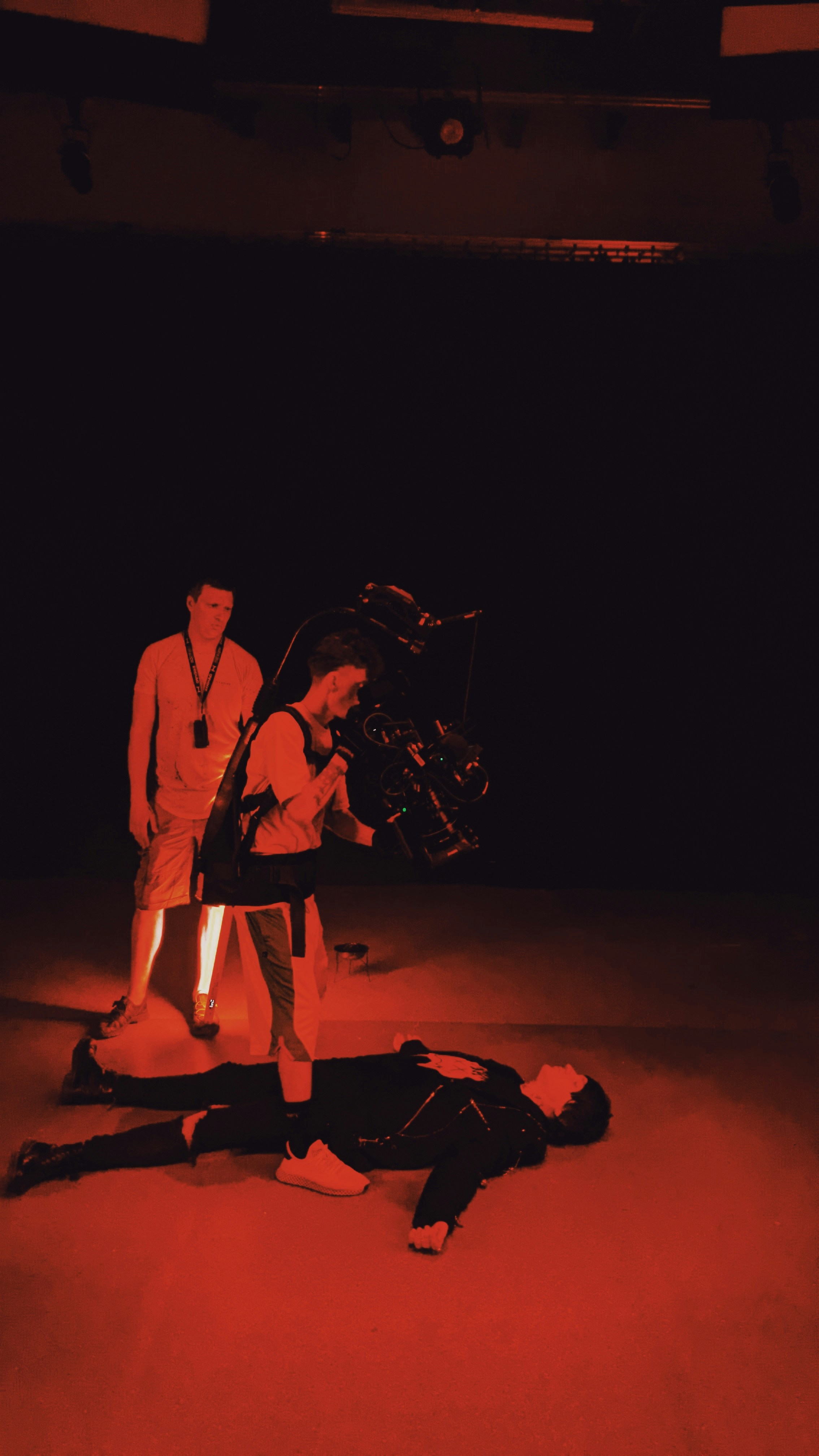 Crew members operate a camera in a dimly lit studio, emphasizing the dramatic red lighting and the subject on the floor.