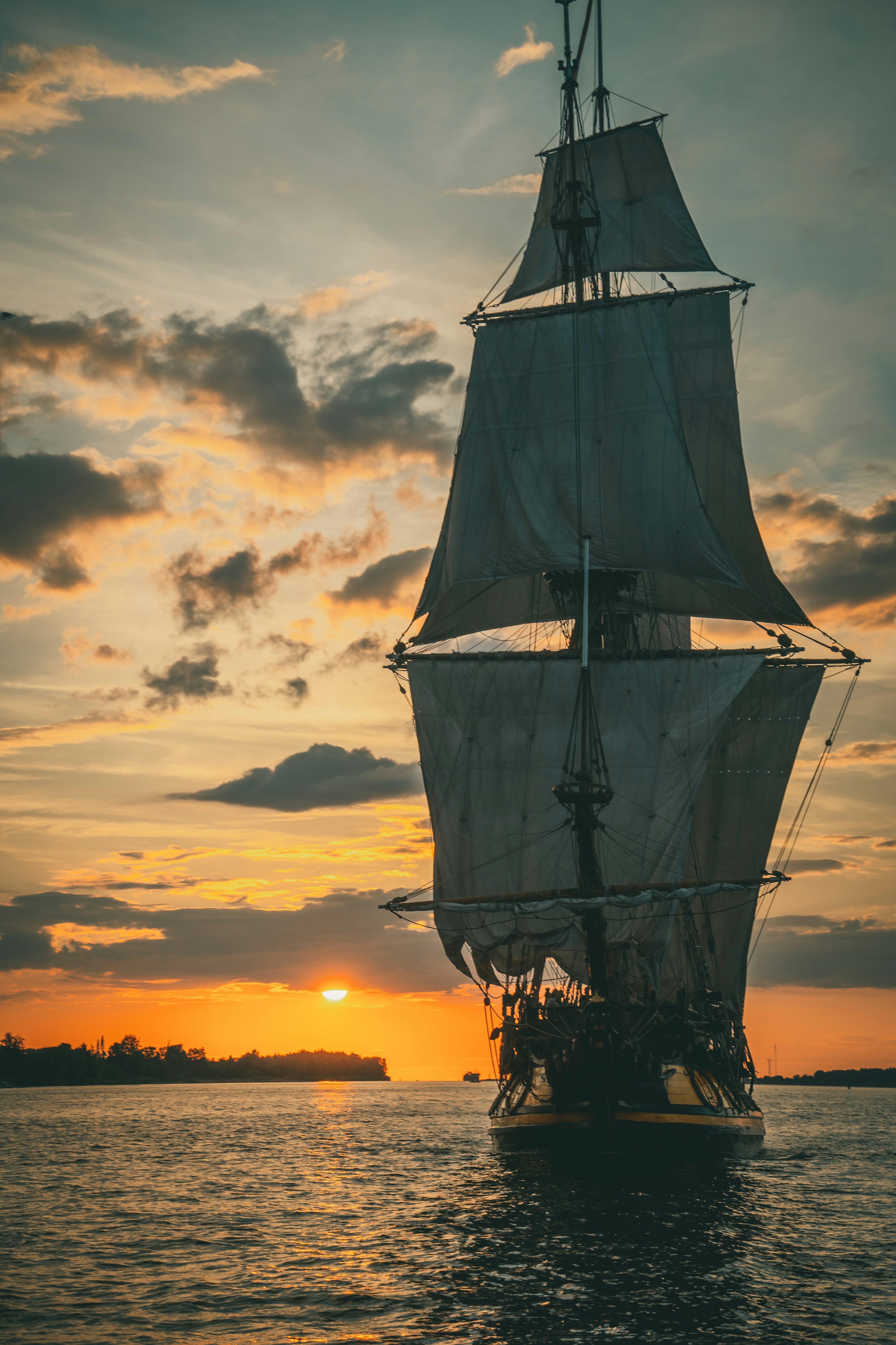 Silhouette of a historical frigate sailing at sunset, with dramatic clouds and vibrant orange hues in the sky.