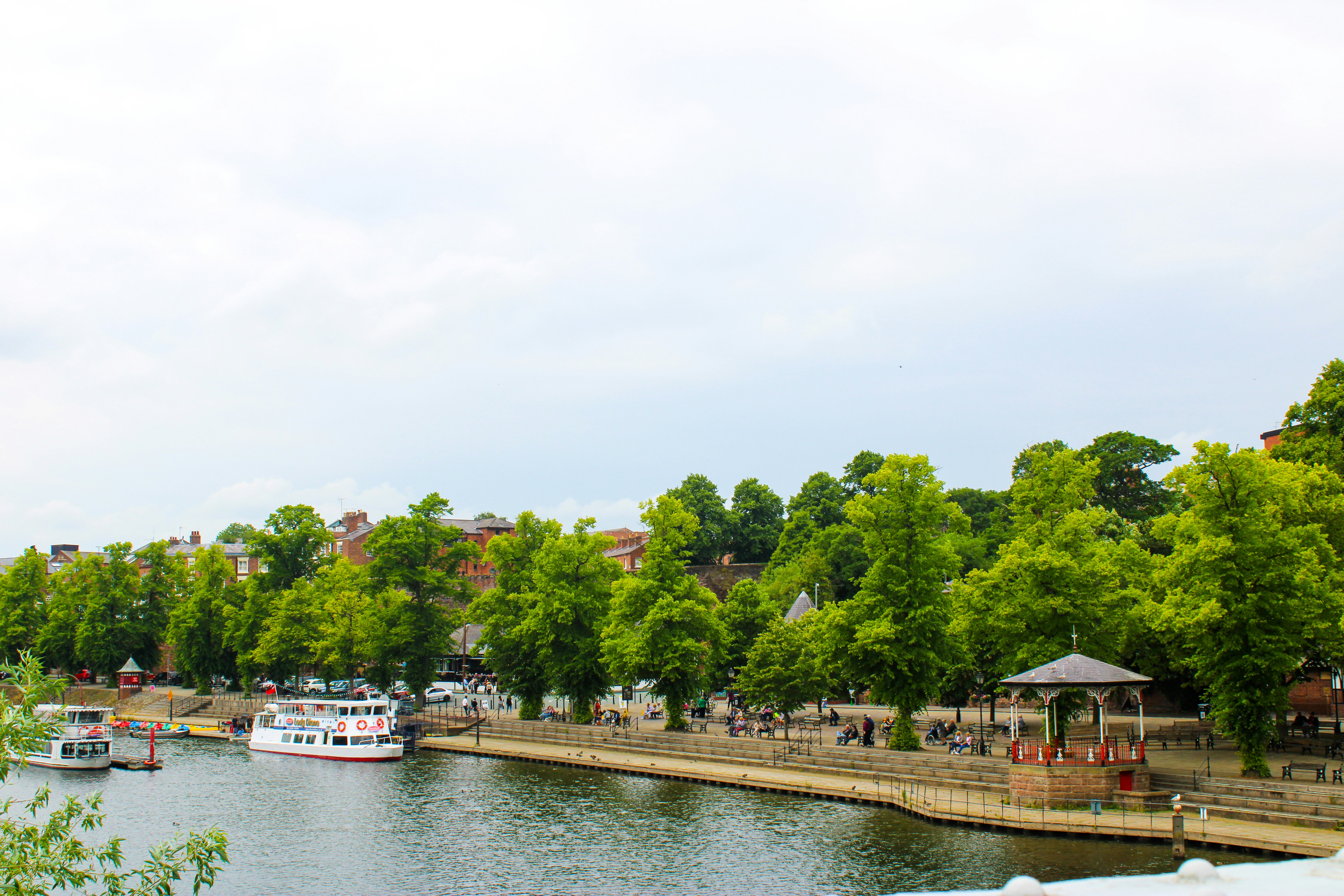 A tree-lined riverbank with boats moored along a calm waterway under a cloudy sky.