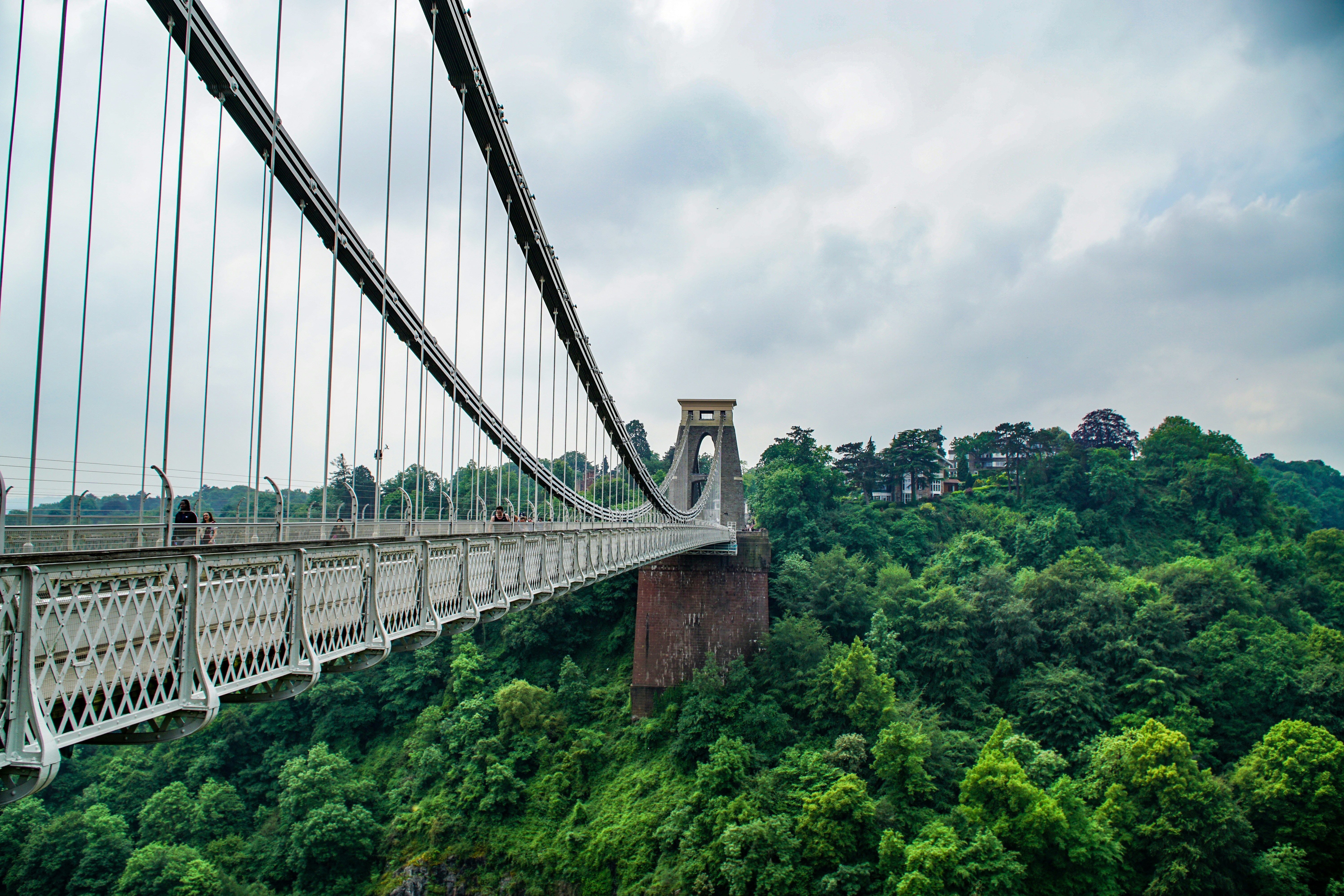 Suspension bridge extends over lush green forest under a cloudy sky.