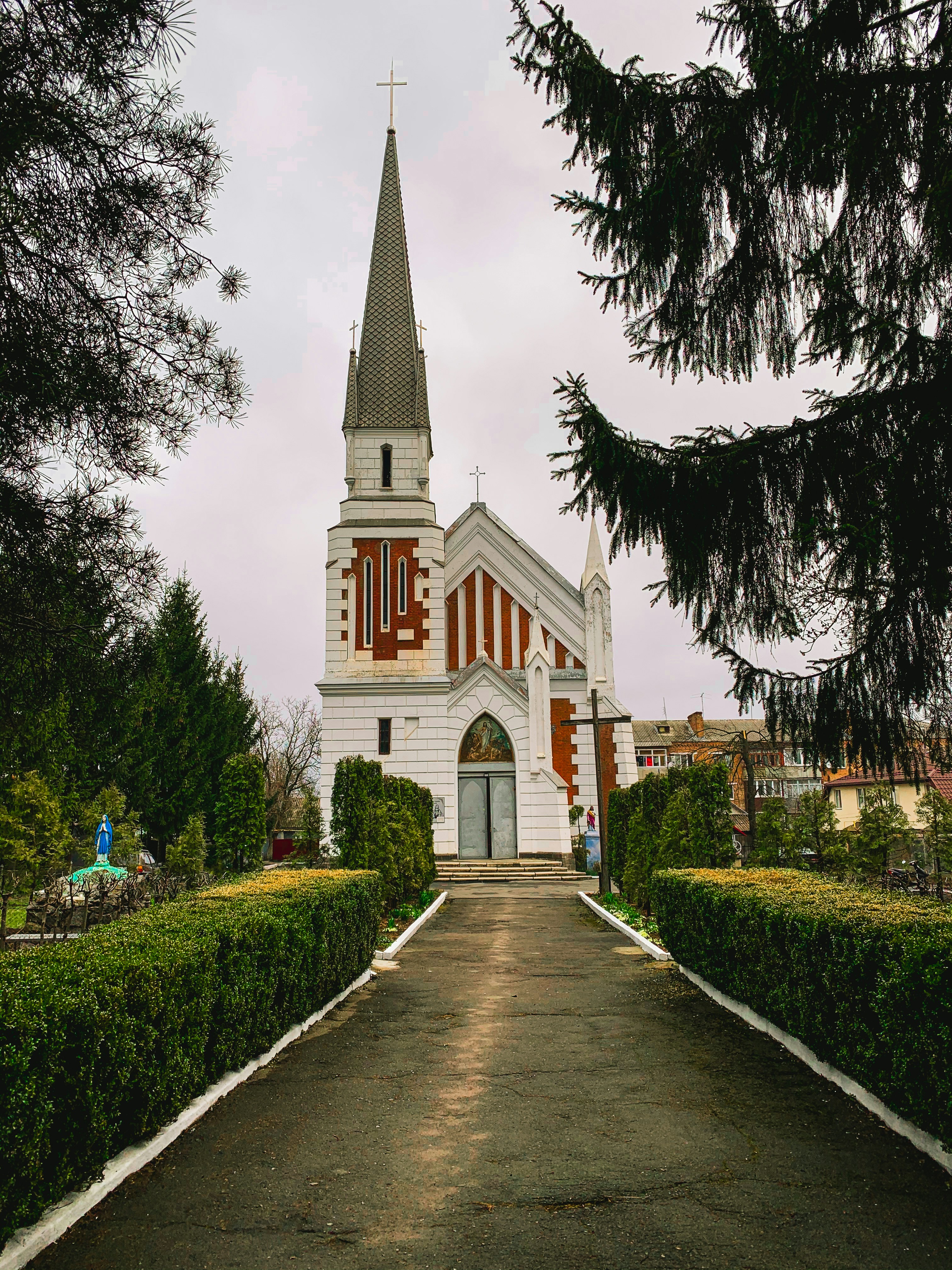 A striking church with a tall spire and distinctive striped facade, framed by neatly trimmed hedges and a pathway leading to its entrance.