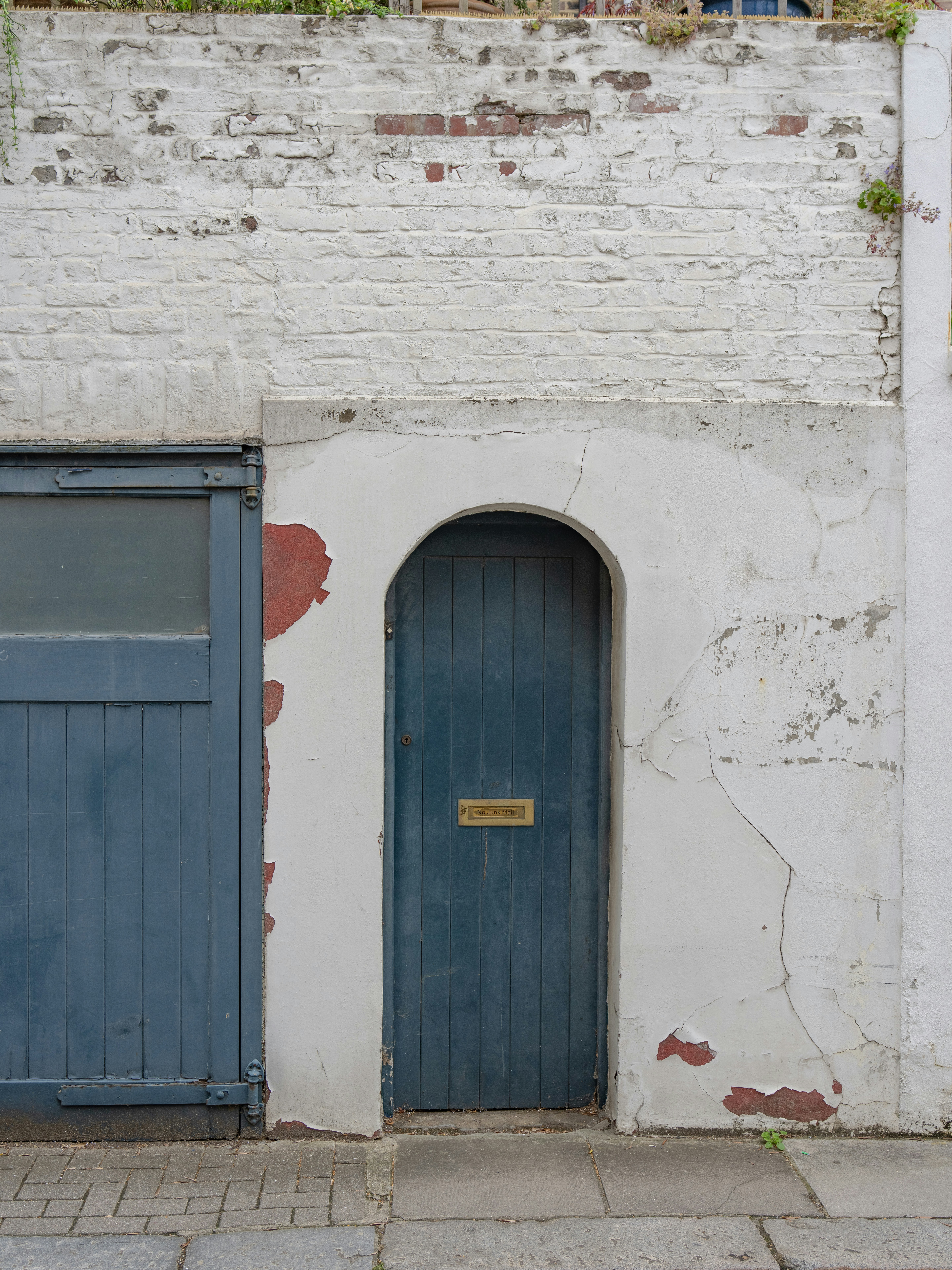 A weathered blue door with an arched top set against a textured white wall, featuring peeling paint and a rustic garage door beside it.