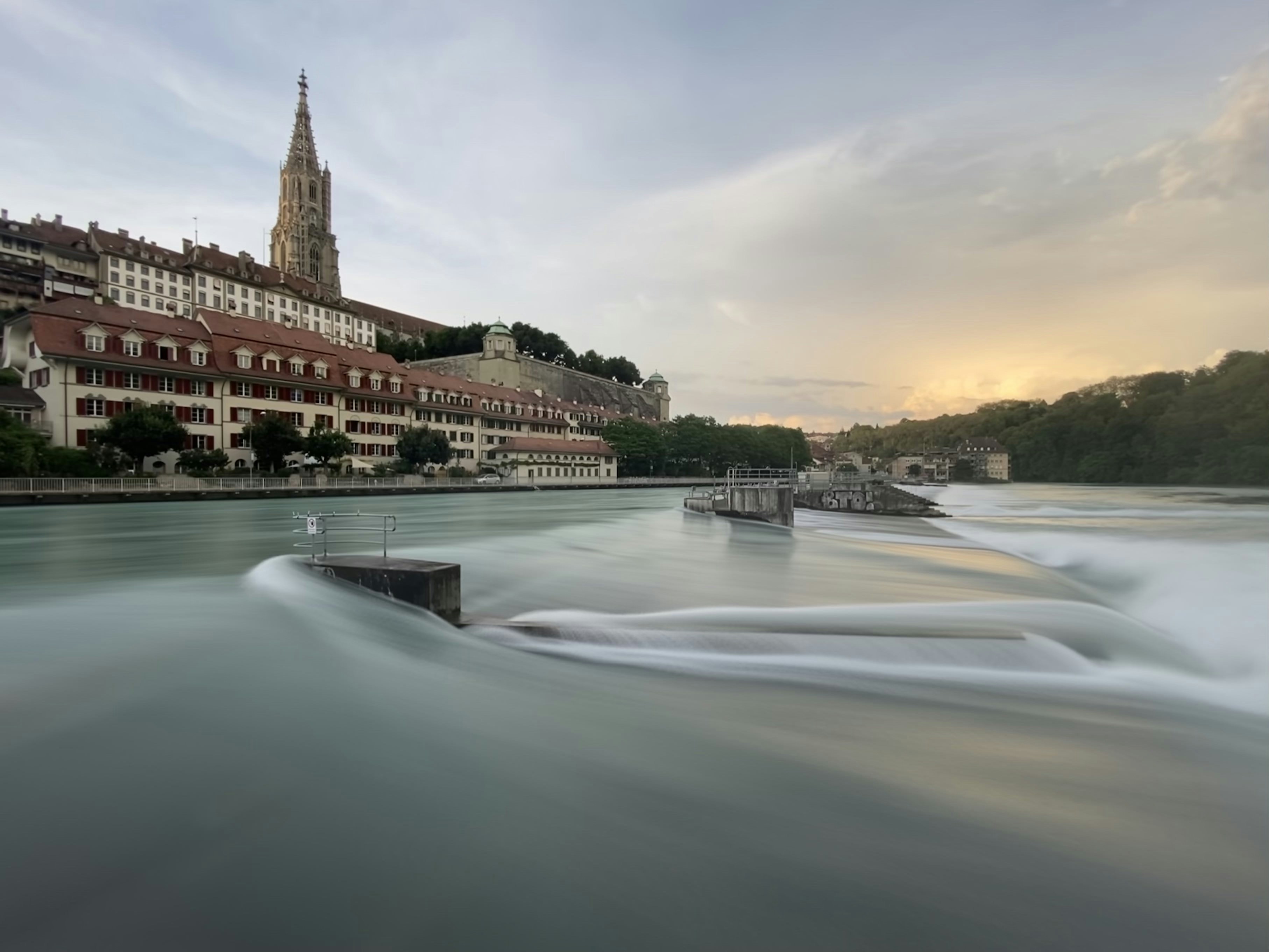 Historic buildings of Bern silhouetted against an approaching storm, with smooth river reflections in the foreground.