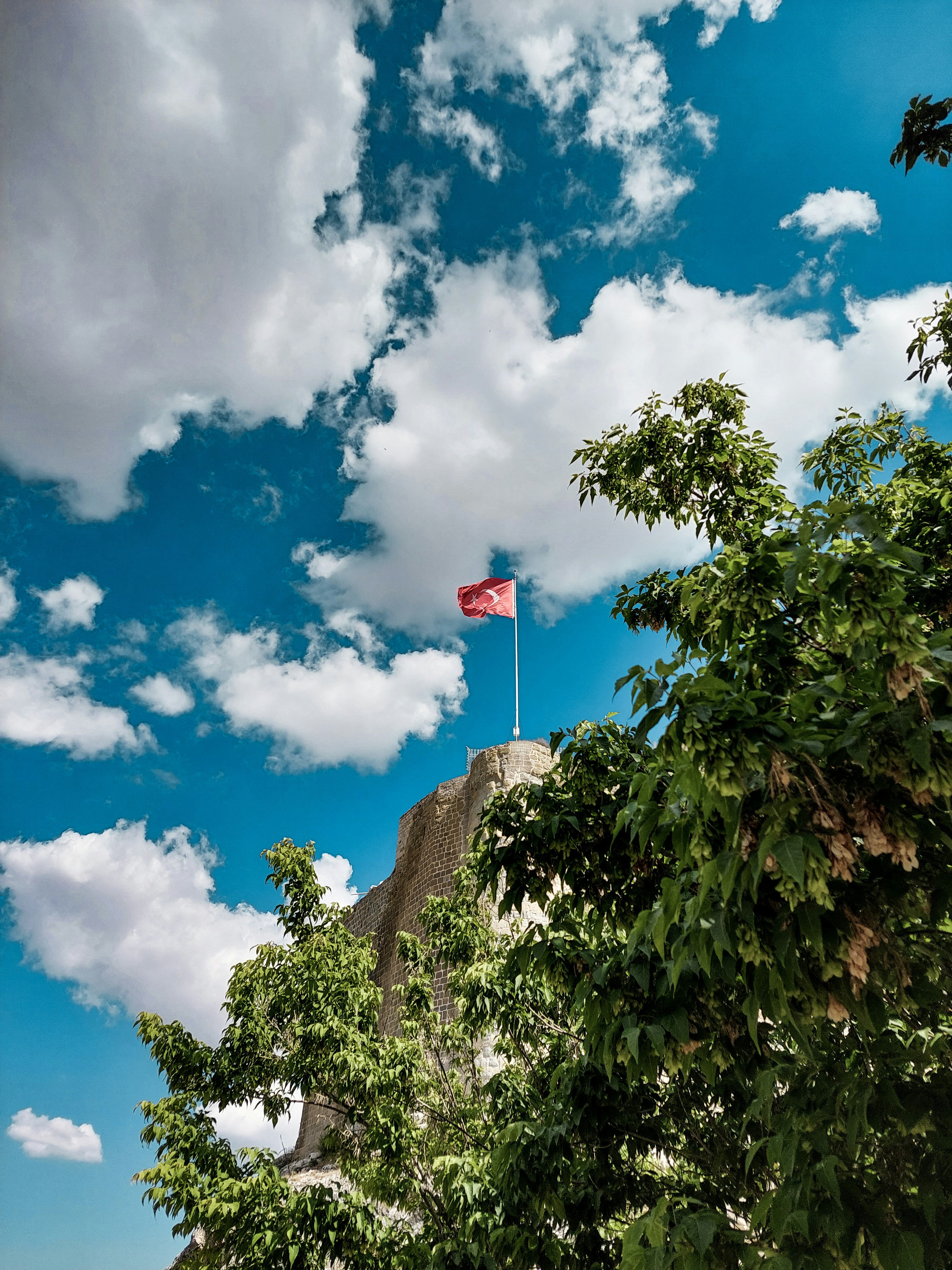 A historic fortress crowned by a Turkish flag, framed by lush green foliage and a vibrant blue sky dotted with clouds.