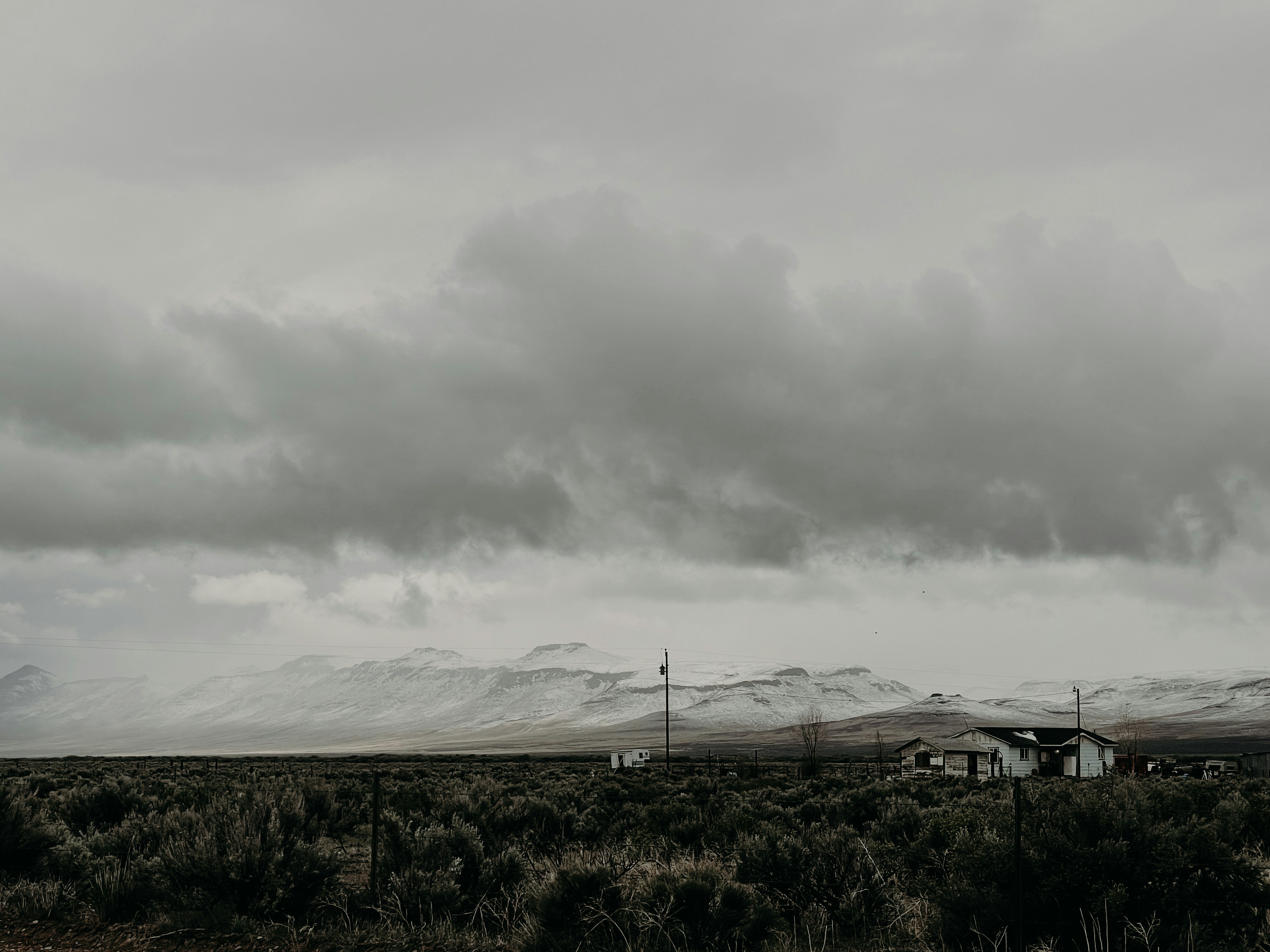 A rustic homestead surrounded by vast sagebrush under a moody, overcast sky. Snow-capped mountains loom in the distance.