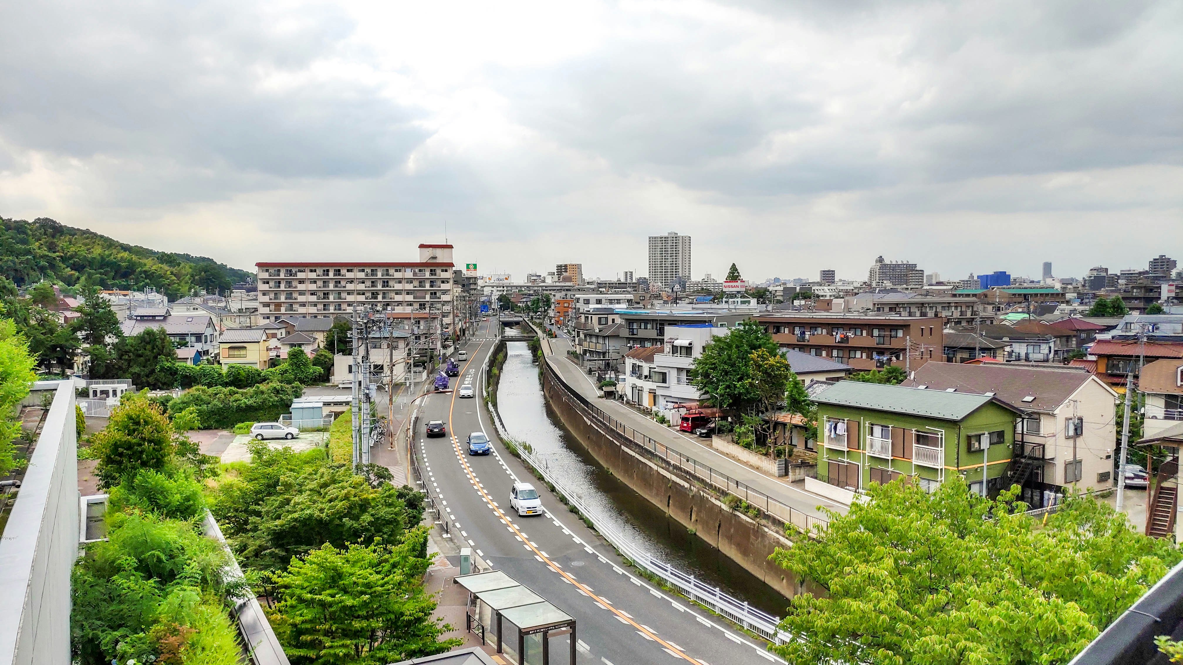 Cars traveling along a curving road surrounded by city buildings and greenery under a cloudy sky.
