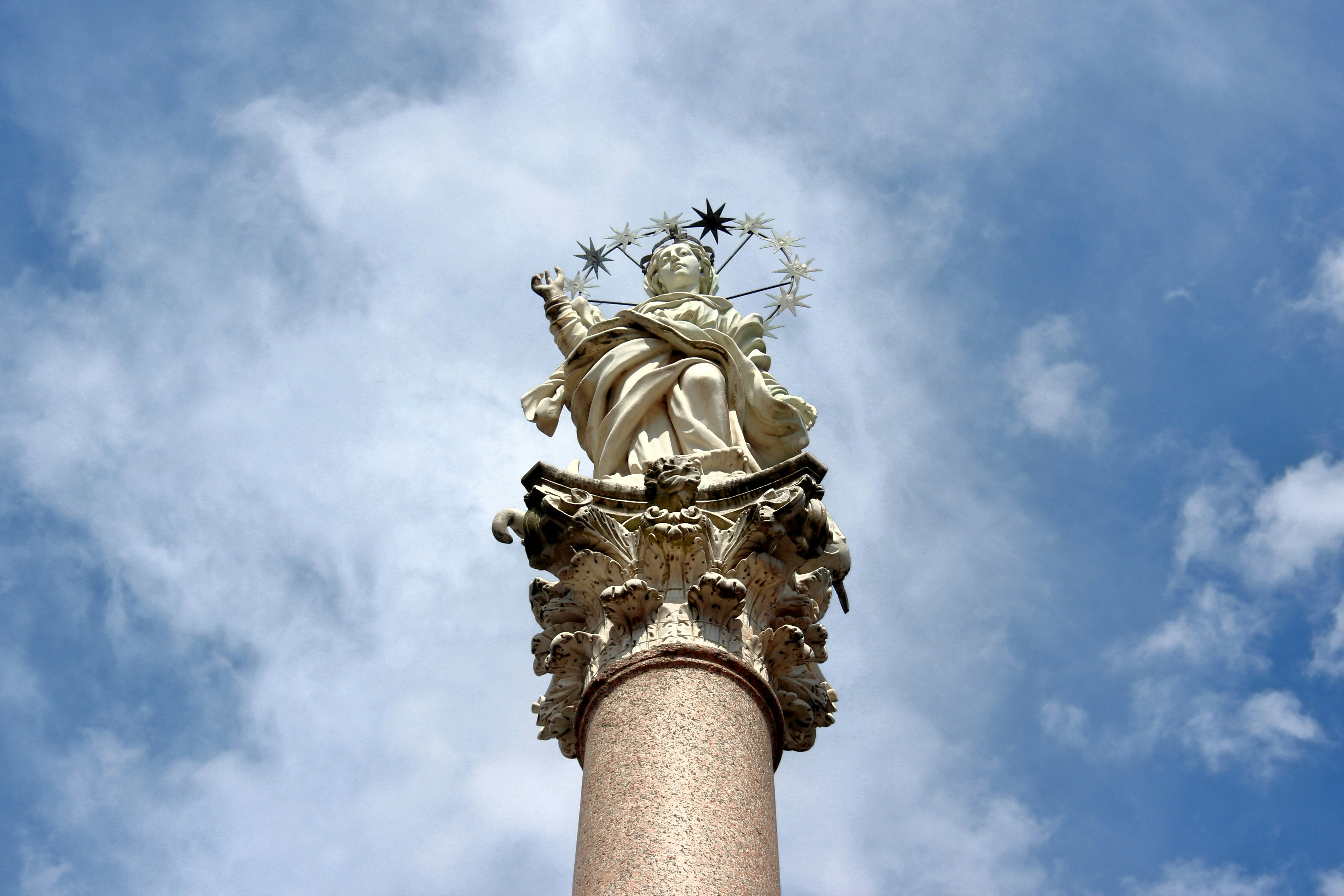 Statue of a figure adorned with stars atop a tall column, set against a blue sky with scattered clouds.