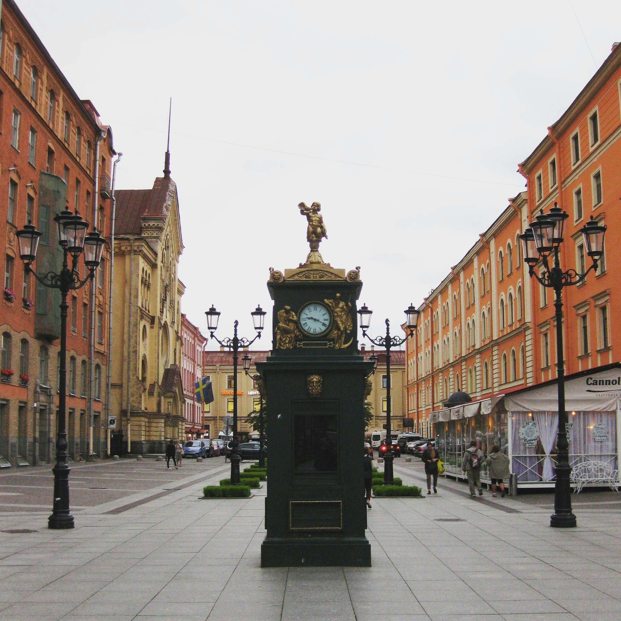 Central clock monument on a paved pedestrian plaza. Pastel-orange historic buildings line the avenue with ornate street lamps.